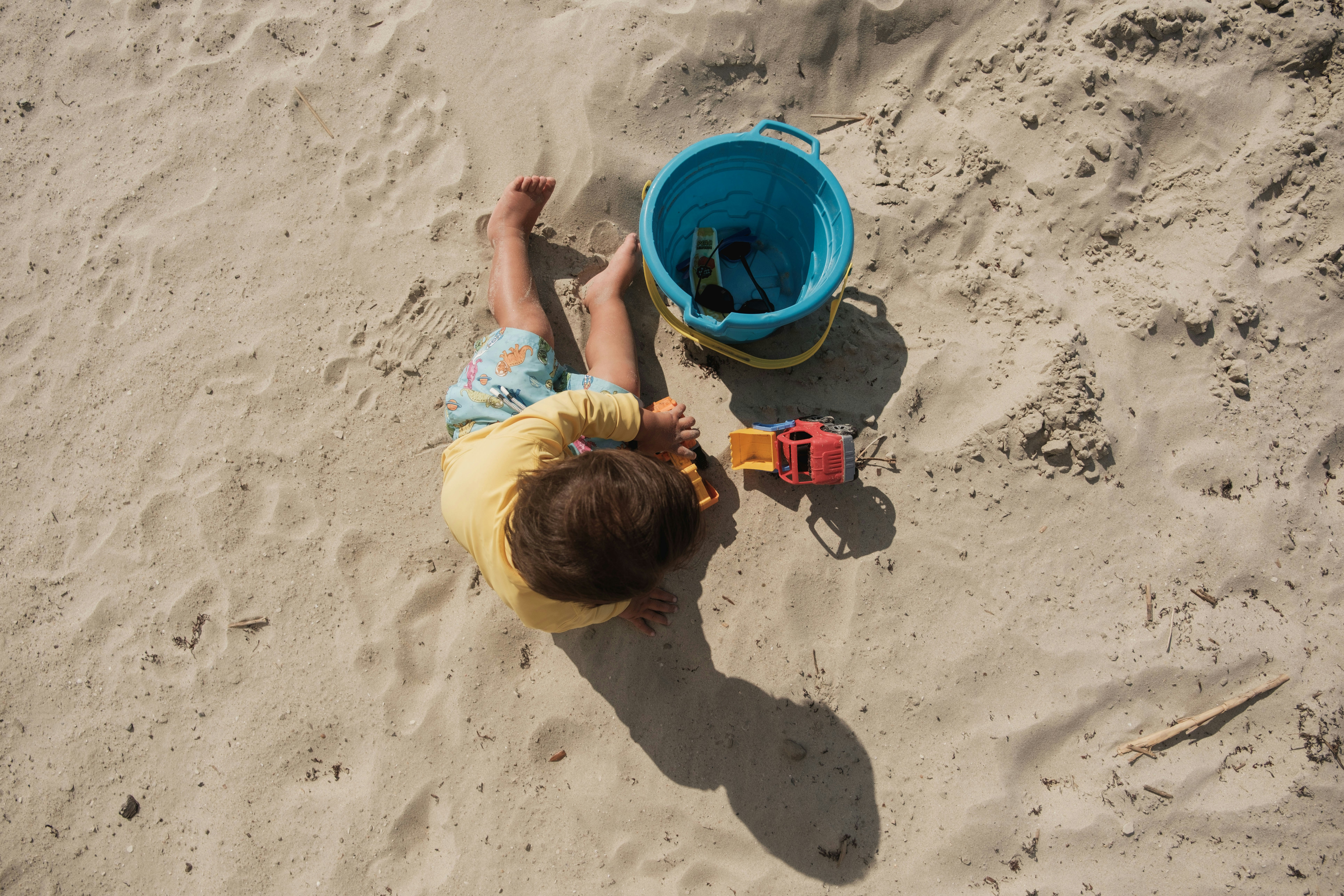 Sandy playground scene with a small child in yellow shirt and blue shorts, a blue bucket, and toy truck from above, capturing a playful moment on the beach-like ground.