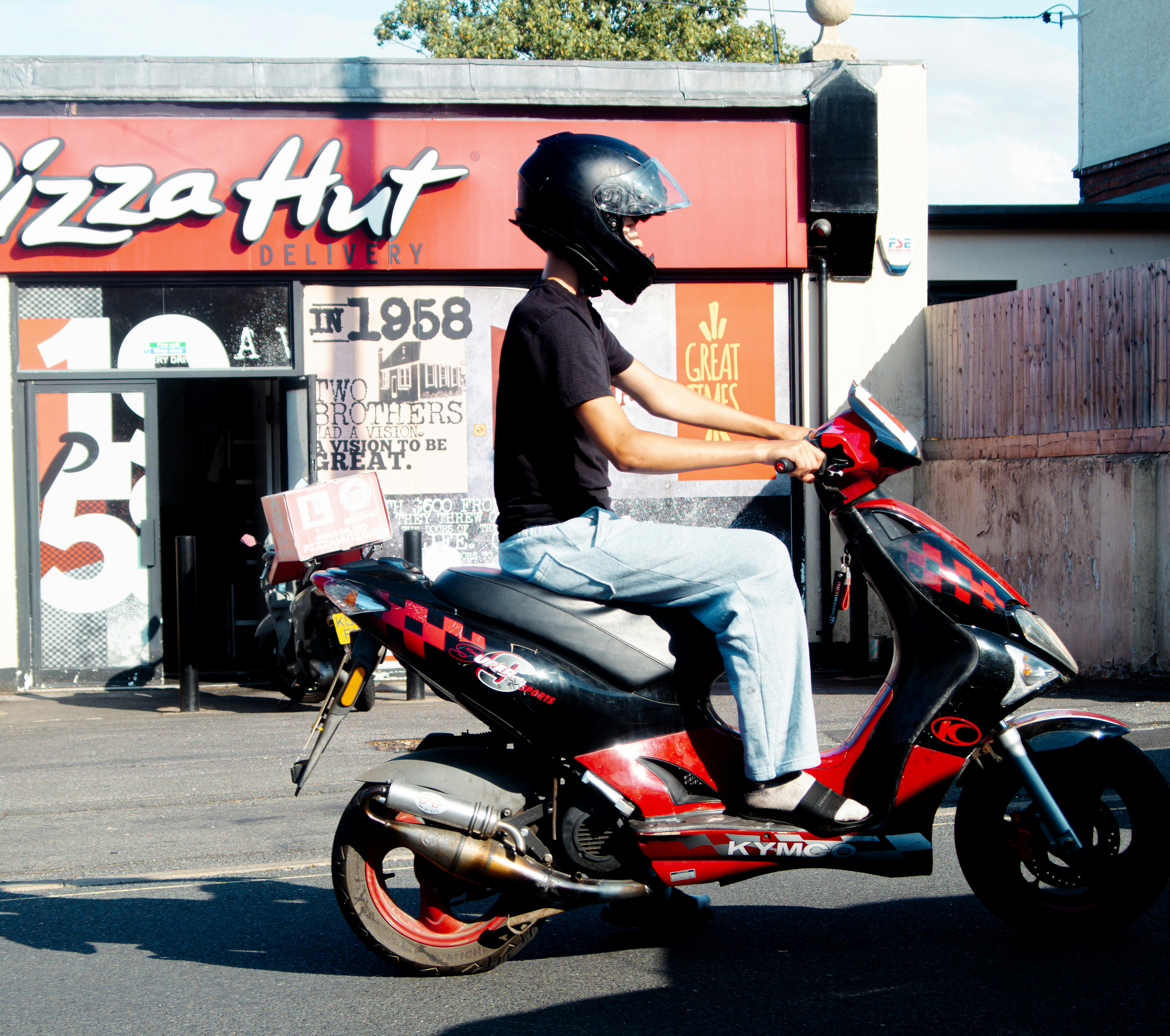 Motor scooter with rider in helmet parked outside a Pizza Hut storefront; bold signage and poster boards create a busy urban scene.
