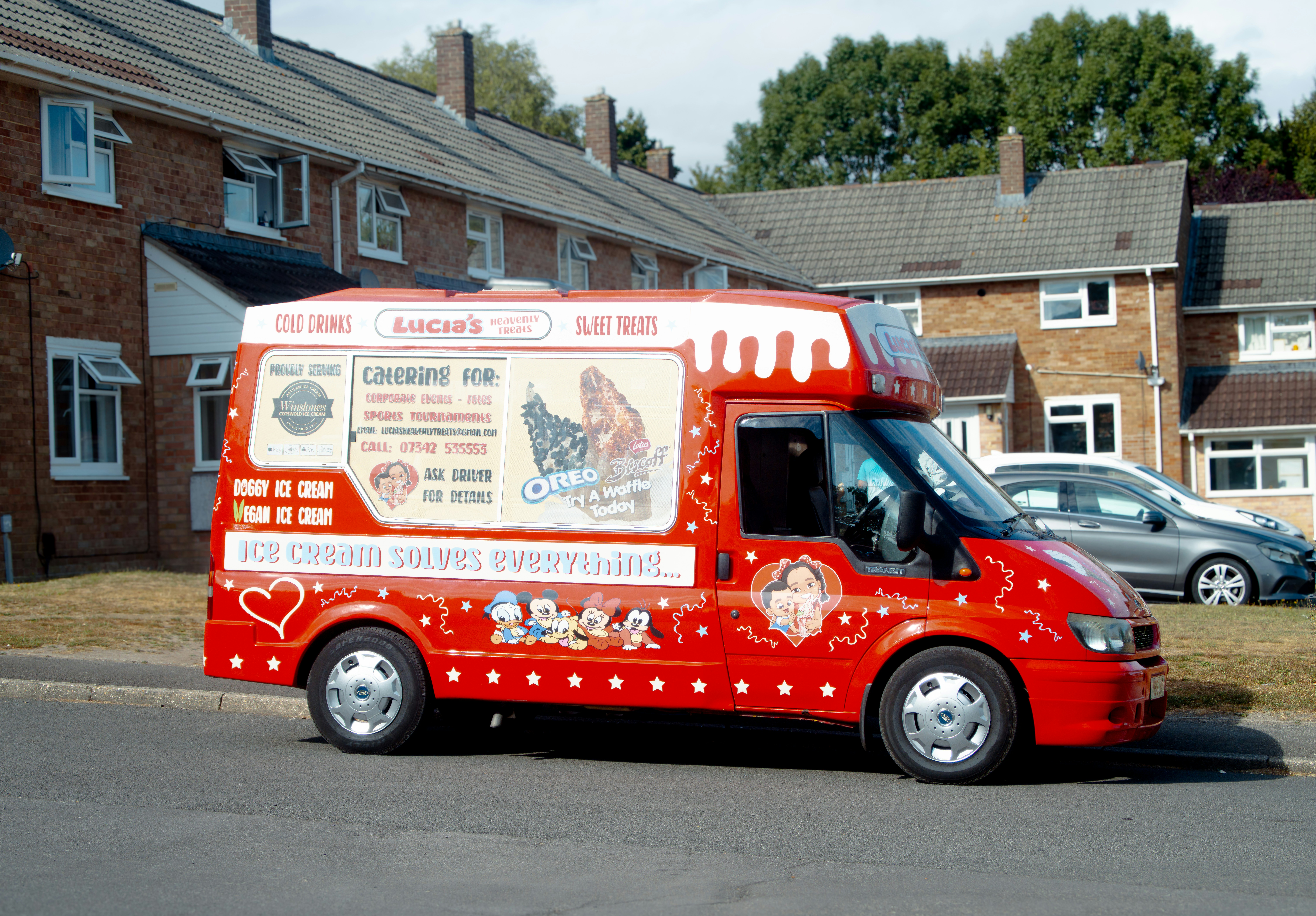 Bright red ice cream van parked along a residential street, with white/cream signage and cartoon graphics. Food truck appears ready for service in a calm neighborhood.