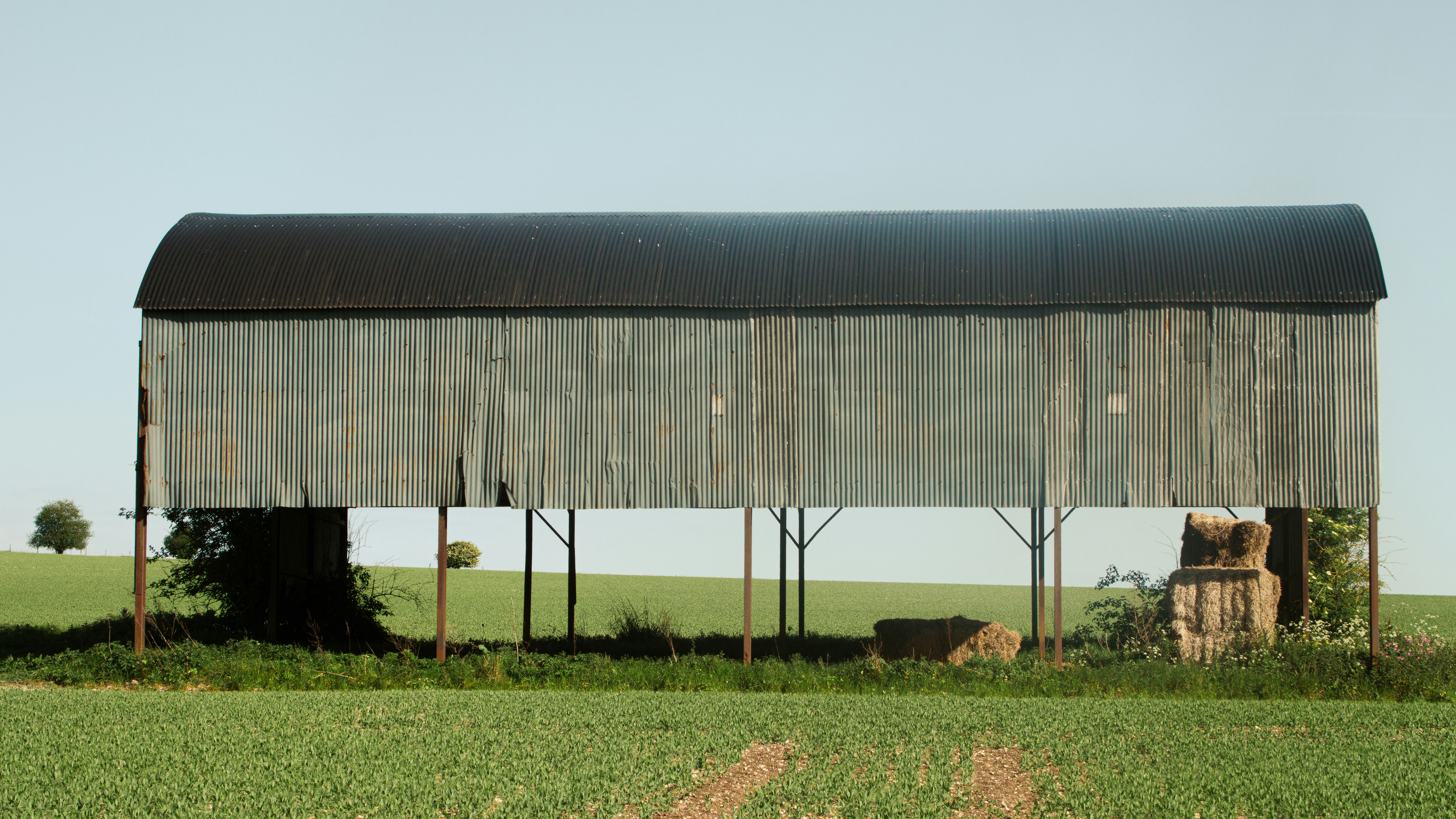 Rusty corrugated shed on stilts with exposed hay bales and open fields beyond, captured in broad daylight.