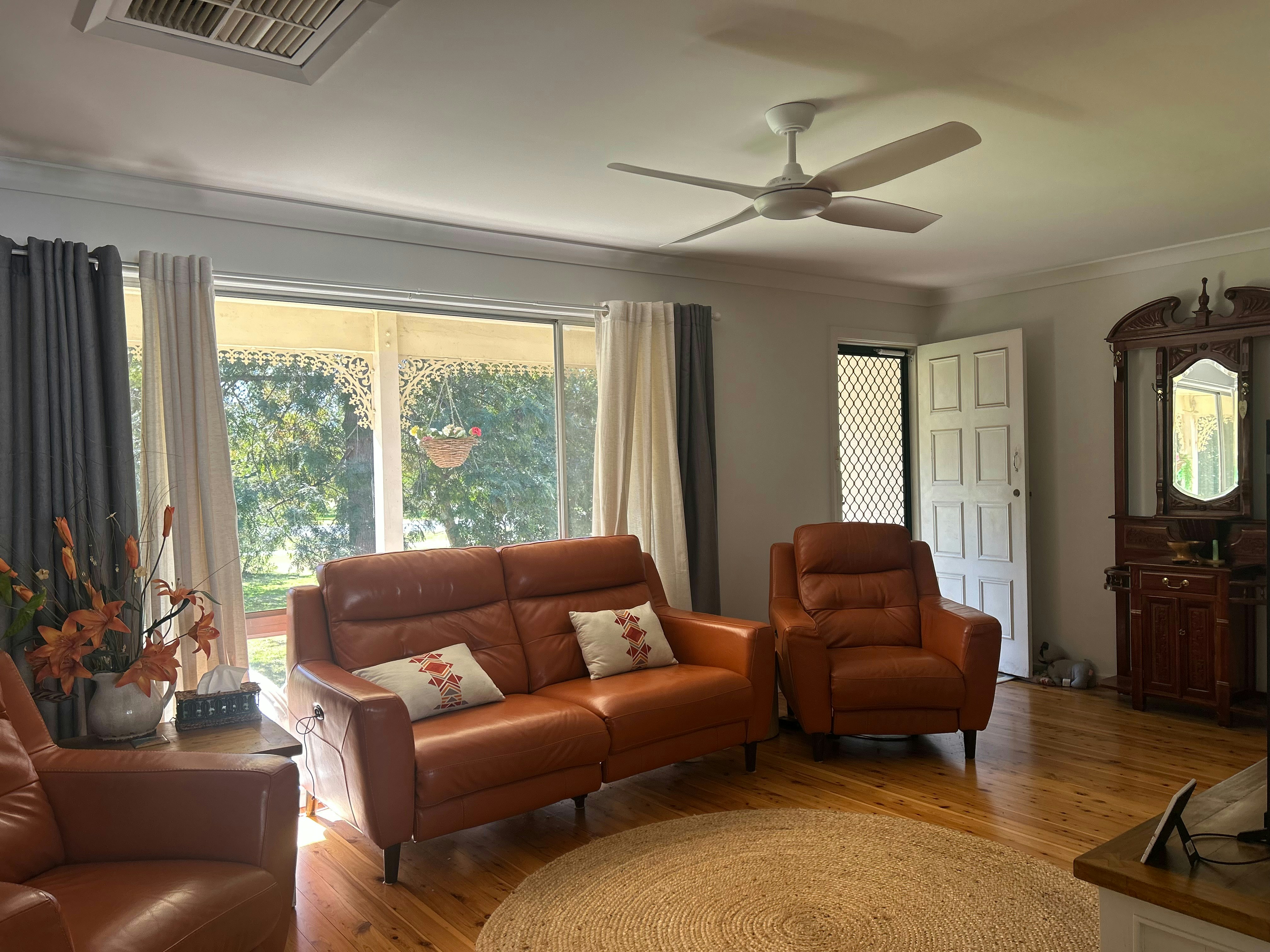 Warm living room with brown leather sofa and armchairs facing a sunlit window; a wooden cabinet and ceiling fan complete the cozy setup.