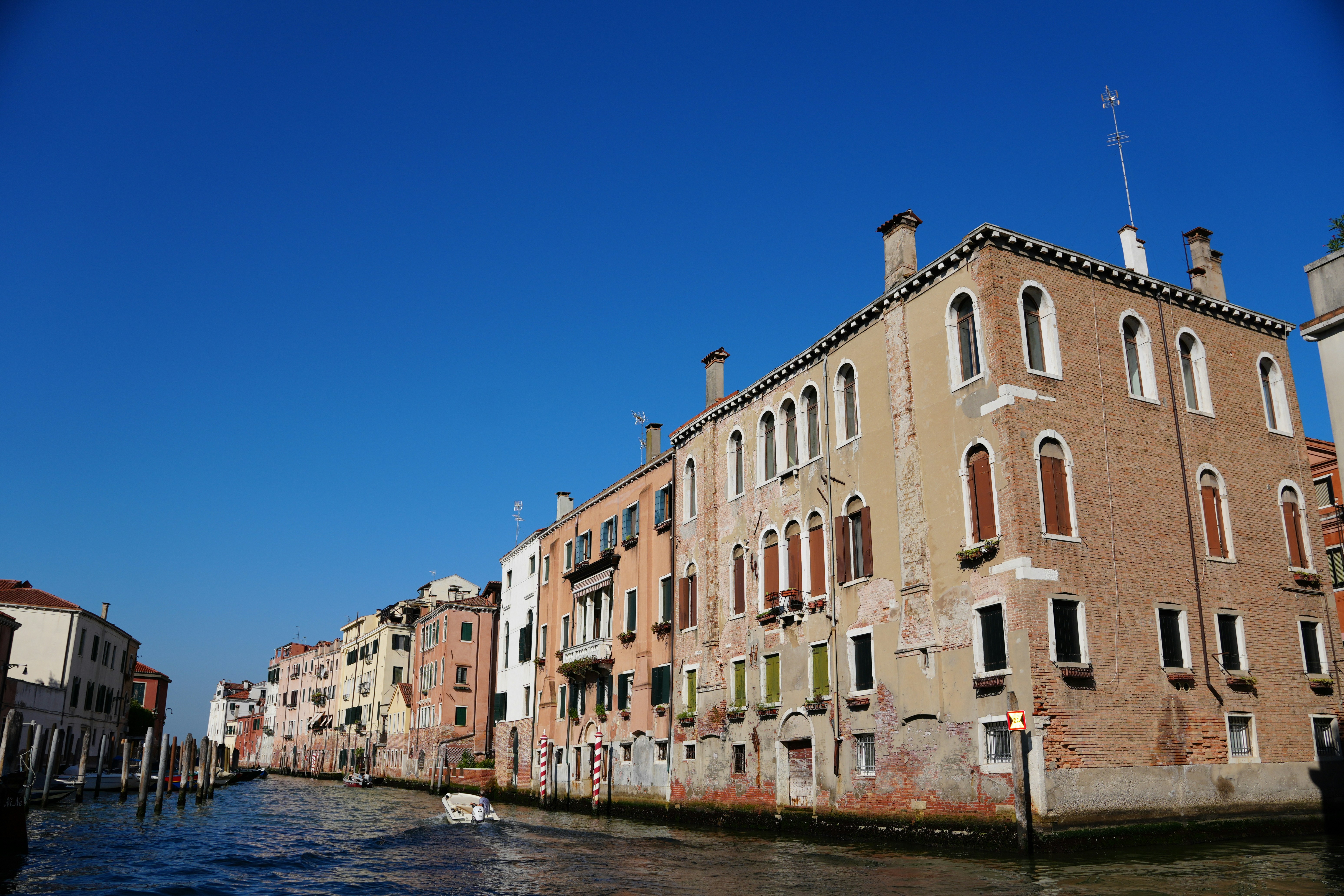 Sunlit Venetian buildings line a calm canal, their weathered facades reflecting in the water while a small boat glides by. A clear blue sky completes the waterfront scene.