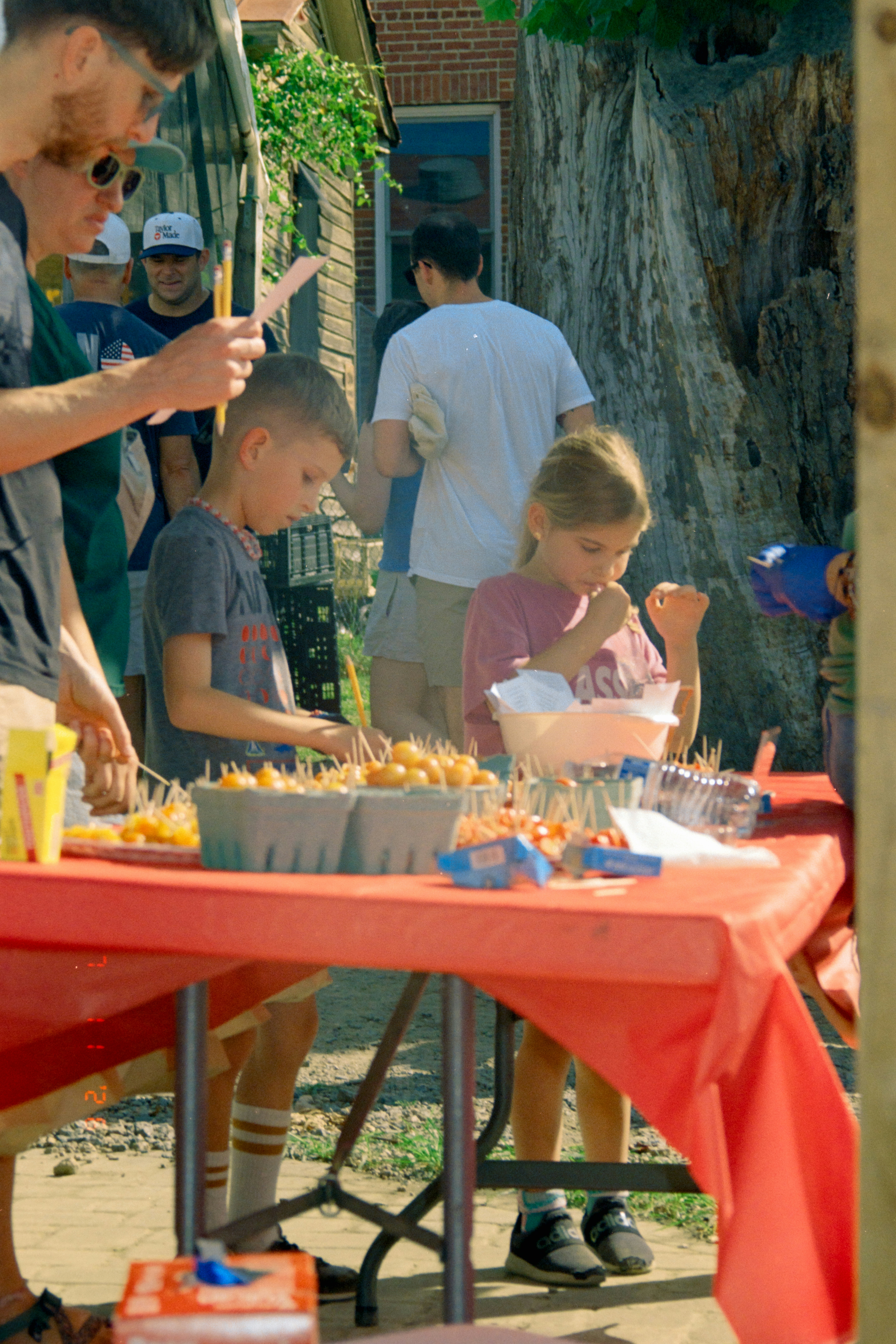 Children at a sunny outdoor snack stall around a red table
