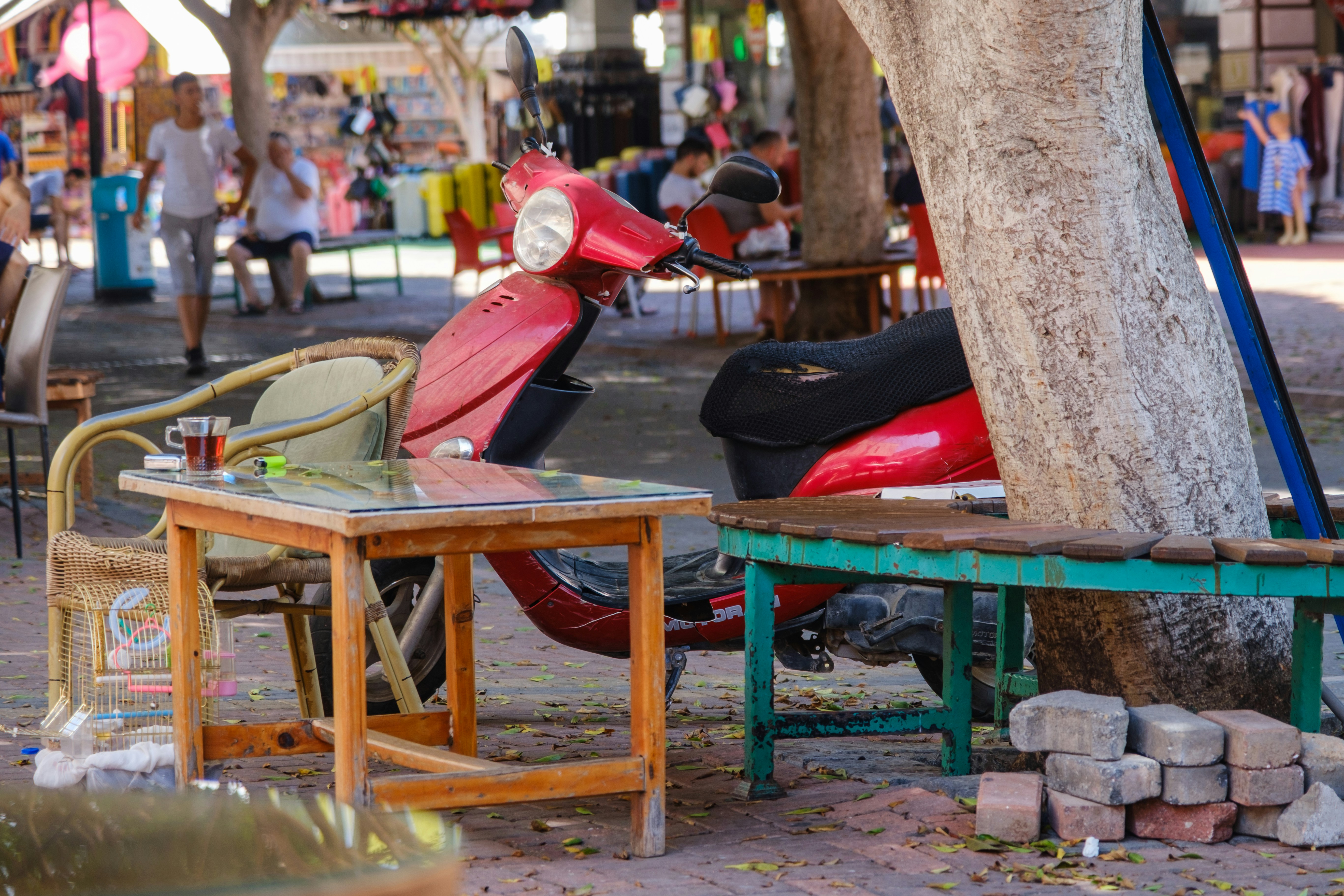 Red Scooter at Rest Under a Splashed Tree in a Busy Plaza
