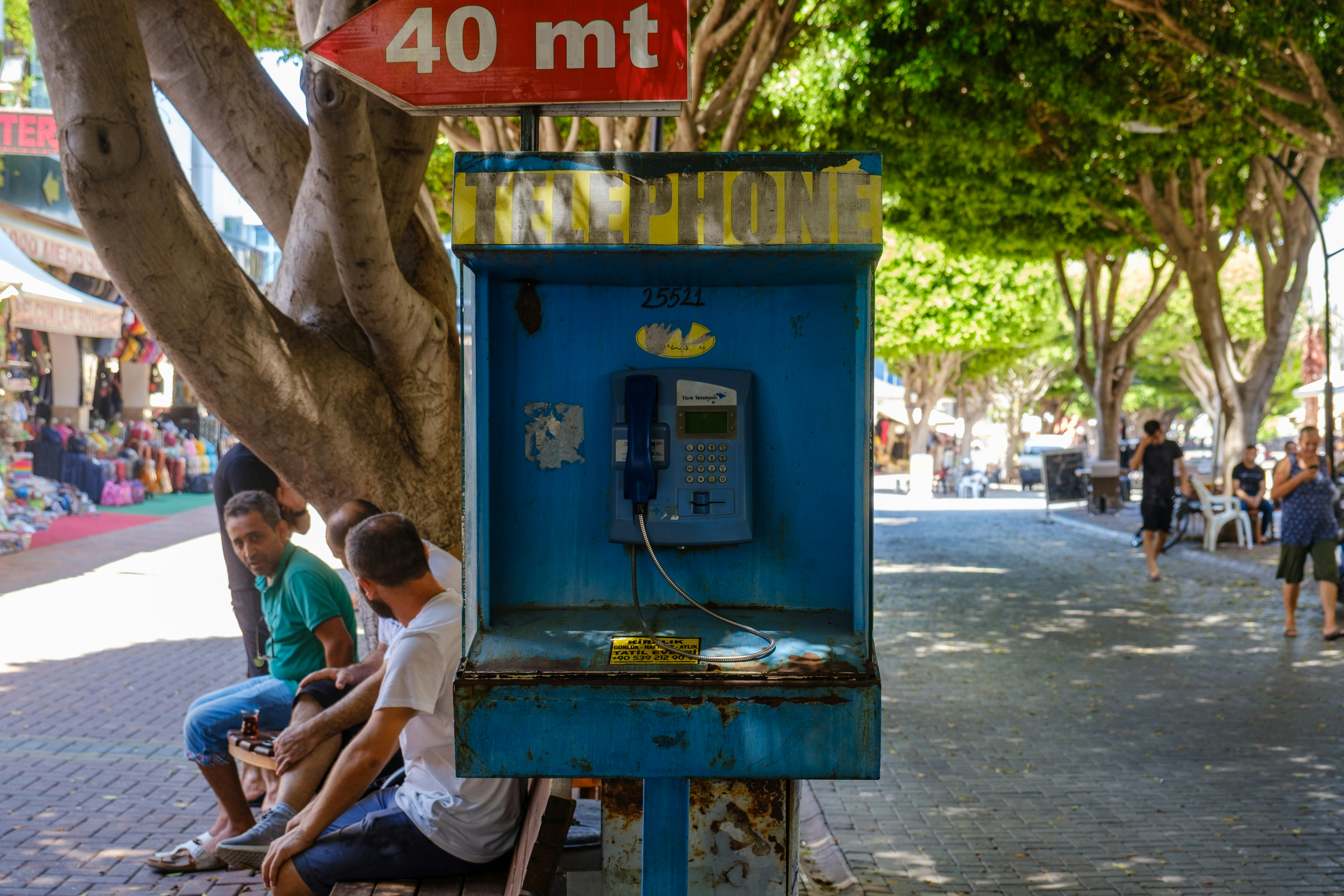 Blue payphone under a leafy canopy in a sunlit market street