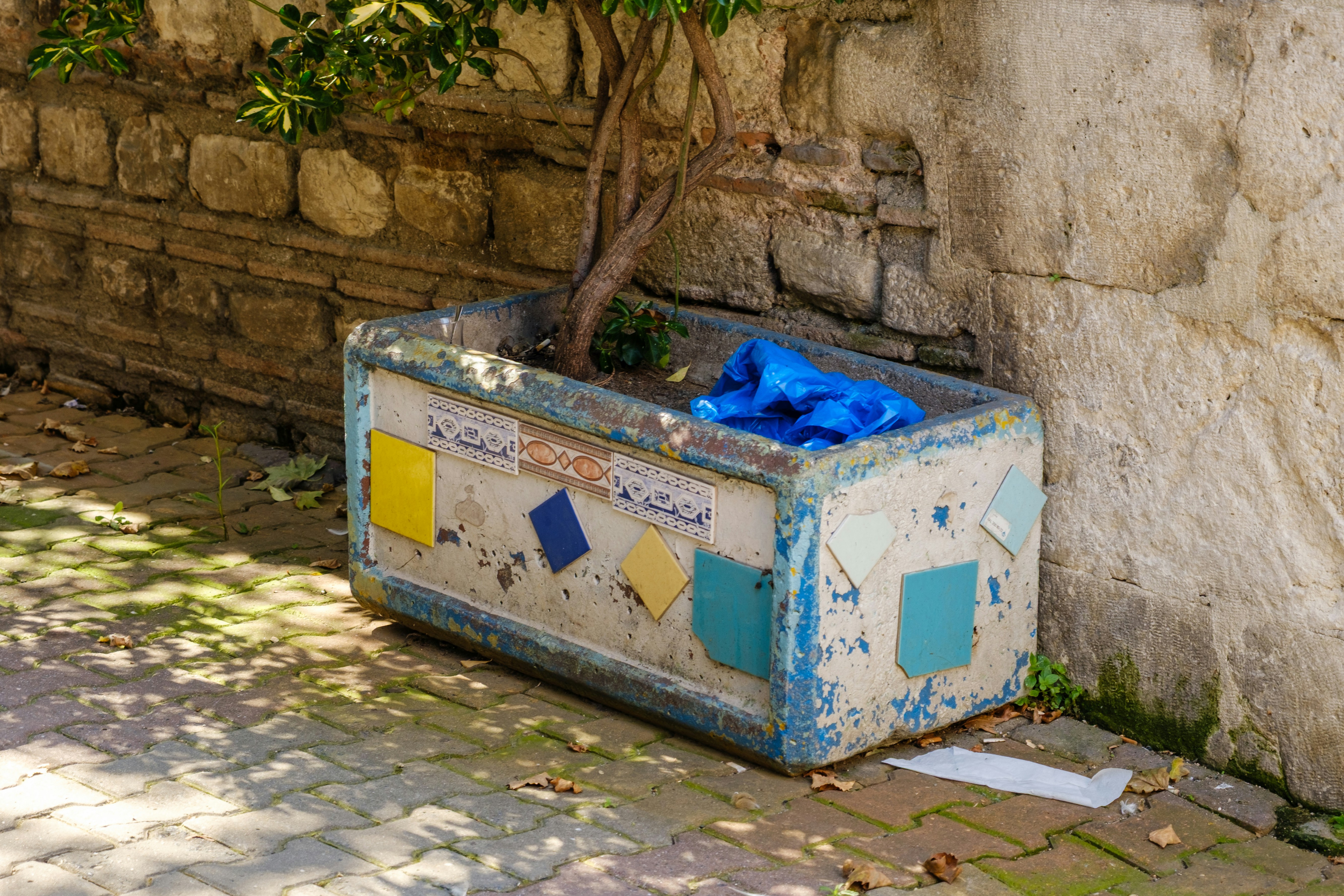 Weathered planter with colorful tiles under a tree by a stone wall