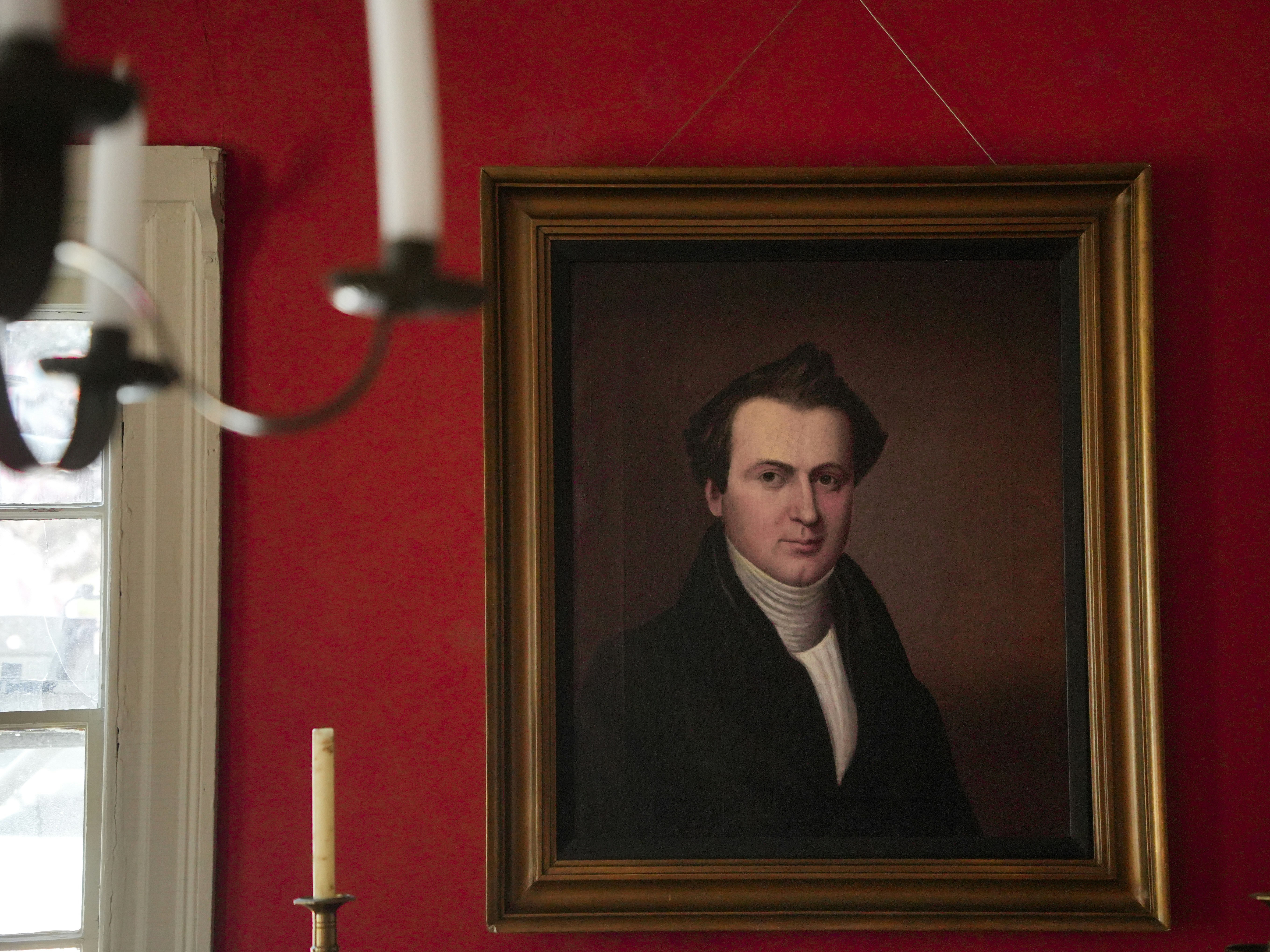 Portrait of a gentleman in a dark coat hanging on a deep red wall; a candle and window are present nearby, suggesting a formal interior scene.