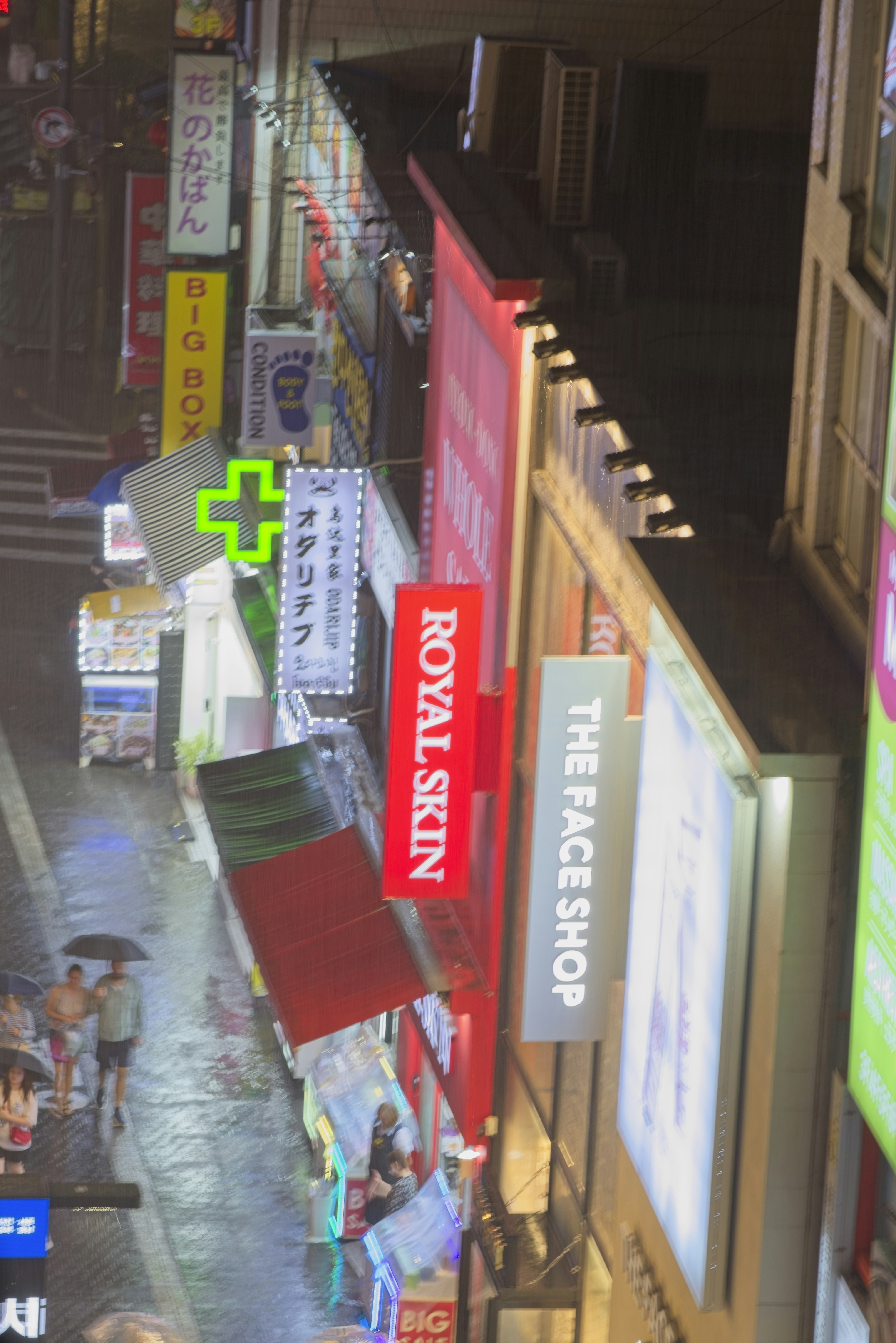 A summer downpour drenches a narrow Seoul shopping street as neon signs blaze against the night — Royal Skin, The Face Shop, and a stack of smaller storefronts glowing in red, yellow, and green. Pedestrians shelter under umbrellas, their reflections shimmering on the rain-slicked pavement below. The humid July evening thickens the air, while overlapping signage creates a dense, electric skyline of commerce. It's a vivid slice of Seoul's after-dark energy, where monsoon rain and neon light combine into a moody, atmospheric urban scene.
