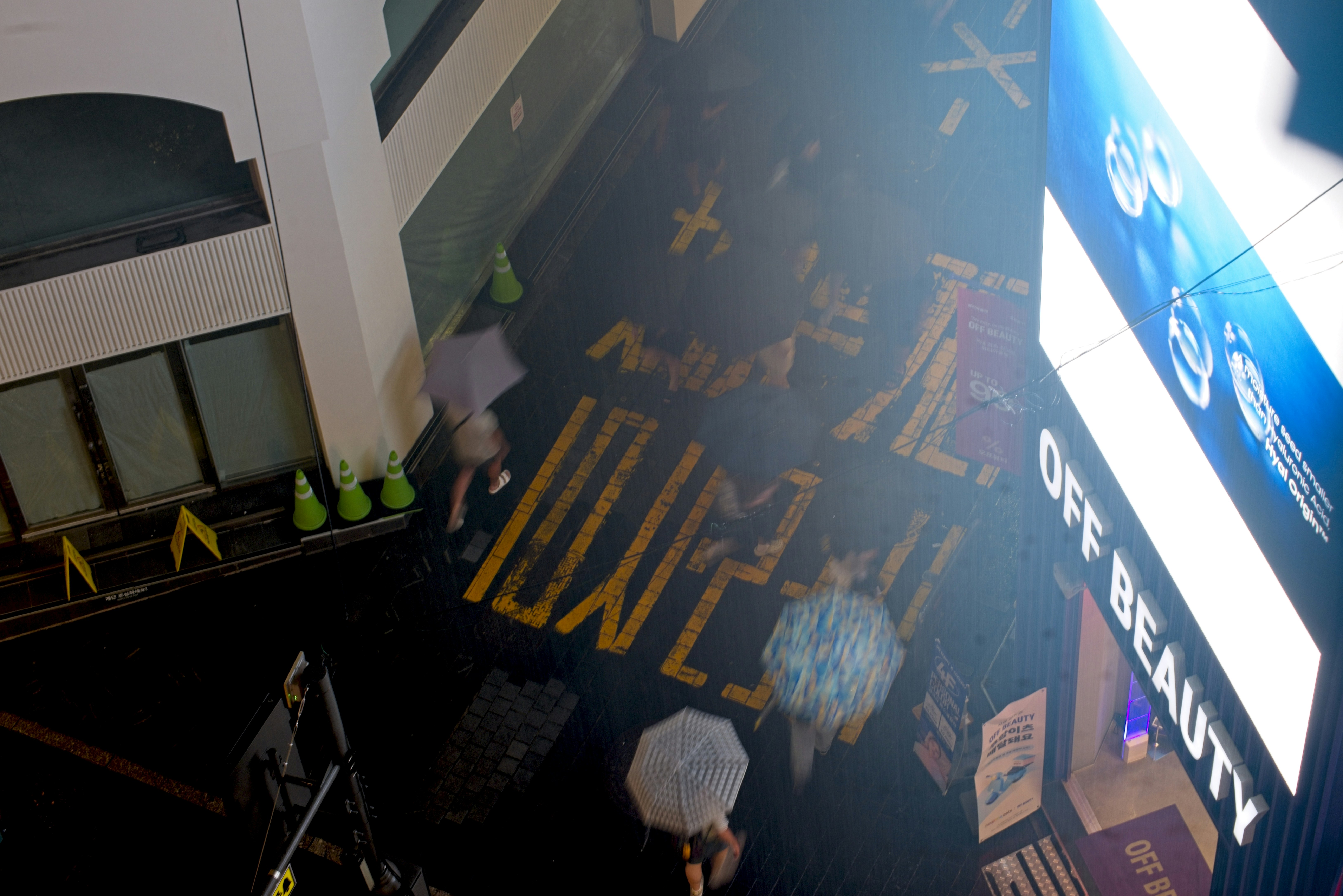 Shot from directly above, this street scene captures pedestrians hurrying beneath umbrellas across rain-darkened pavement marked with bold yellow Korean lettering. Motion blur from the downpour softens the figures below, while green traffic cones and warning signs dot the sidewalk's edge. To the right, the illuminated "OFF BEAUTY" storefront glows against the wet night, its cosmetics ads reflecting on the slick tiles. The bird's-eye angle and streaking rain create an almost abstract, cinematic composition — a striking, moody capture of a humid July evening downpour in downtown Seoul.