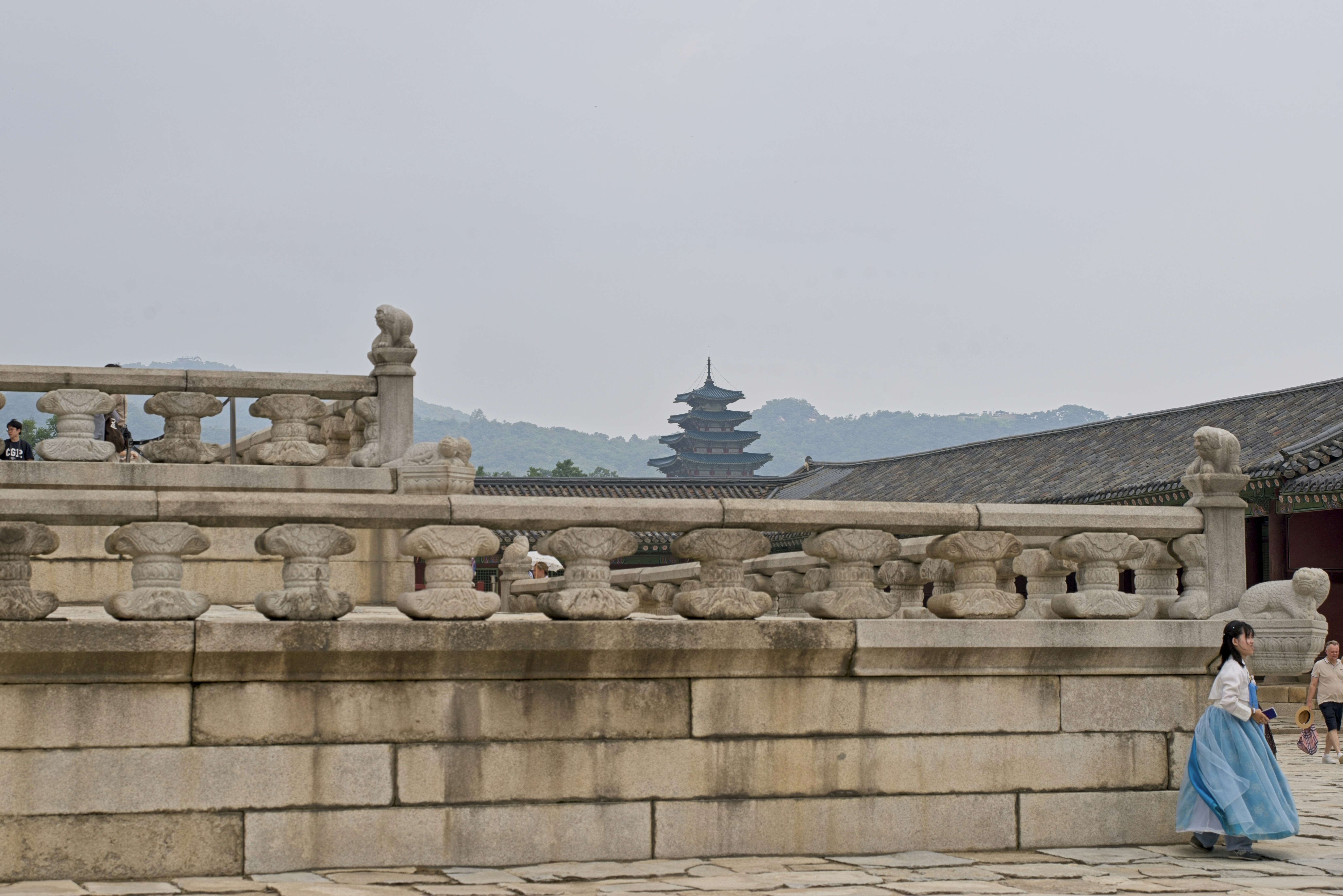 Stone railing and platforms frame a distant pagoda while a person in a blue hanbok-like dress stands near the right foreground; the scene resembles a traditional palace setting with soft daylight.