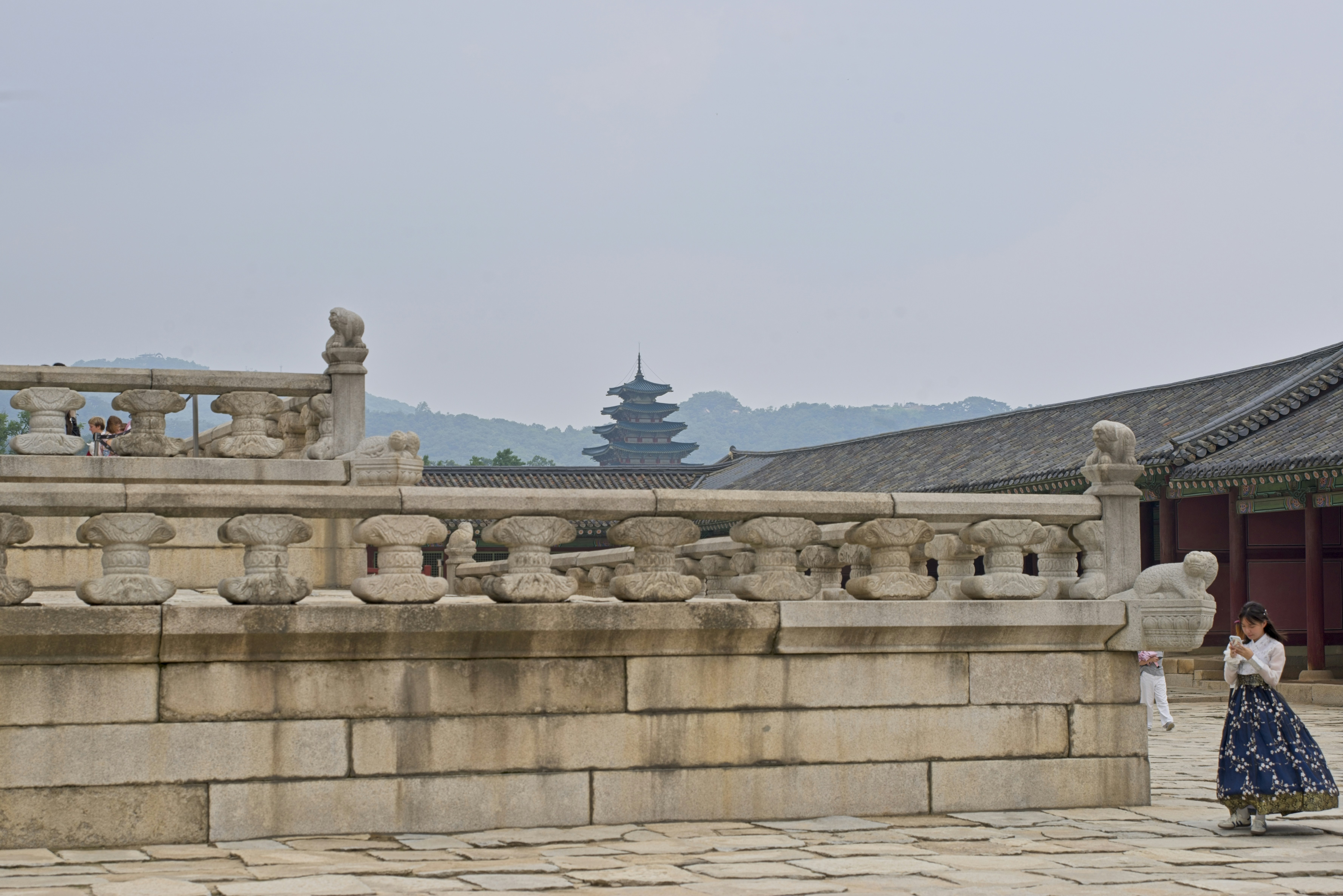 A woman in a navy floral hanbok checks her phone beside an ornately carved stone balustrade at Gyeongbokgung Palace, its guardian lion statues standing watch at the corners. In the hazy distance, Hyangwonjeong pavilion's tiered roof rises above palace rooftops, framed by soft green mountains. Other visitors linger along the terrace above, taking in the view. The weathered granite railings showcase intricate lotus-like carvings, adding texture to the foreground. A quiet, contemplative moment set against the grandeur of Seoul's royal architecture on a hazy summer day.
