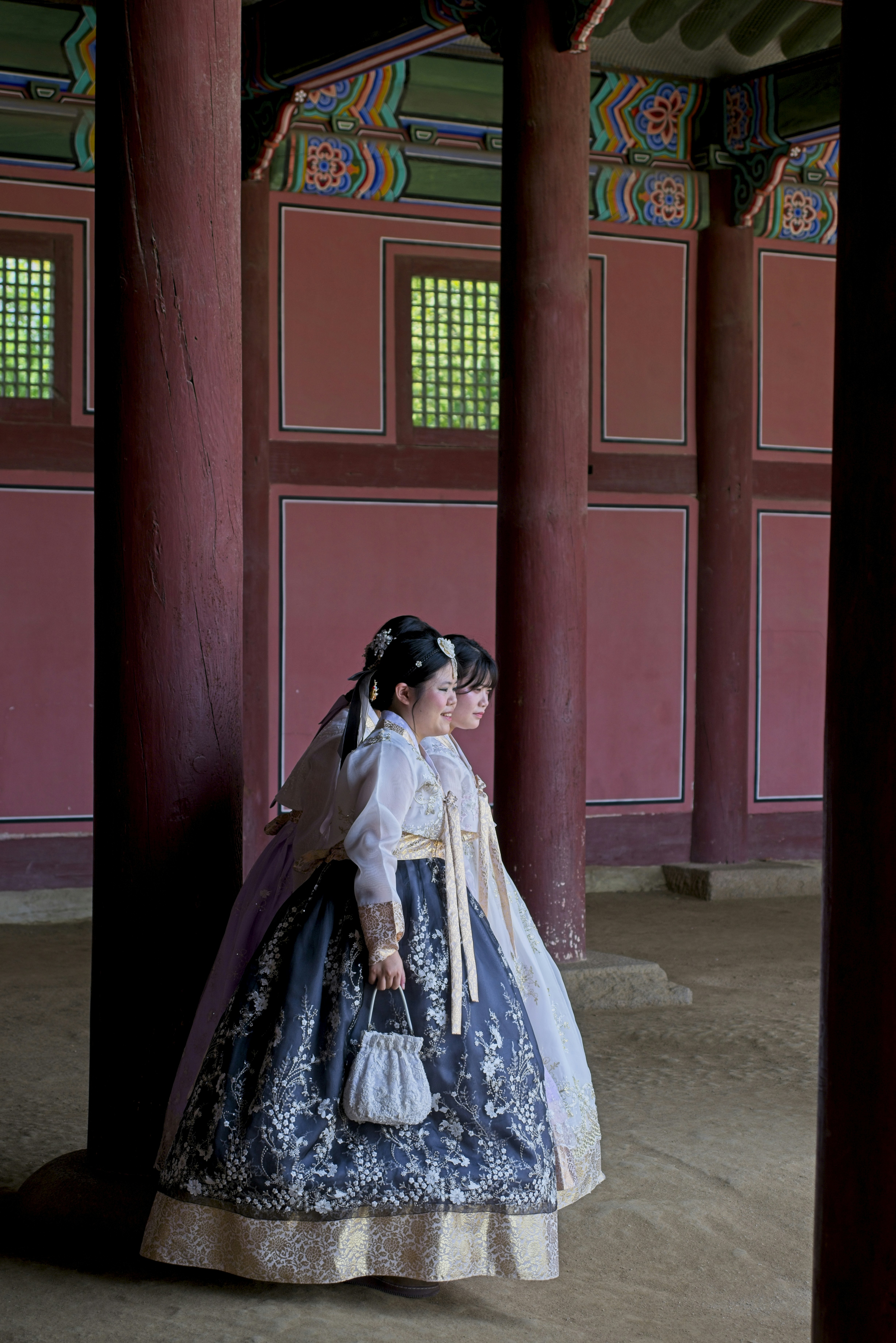 Two Traditional Hanbok Wearers Walking Between Crimson Columns
