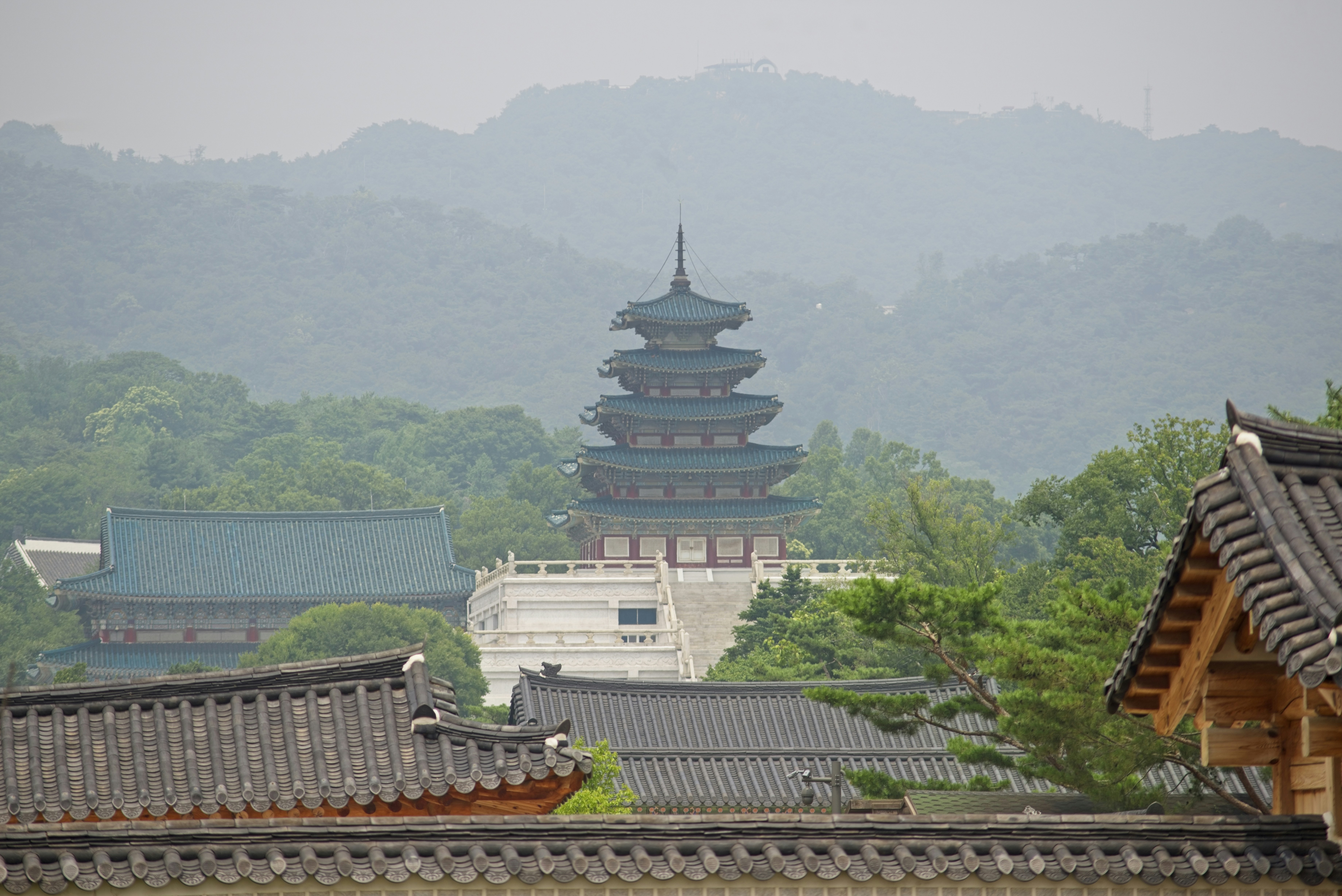 Layers of traditional Korean rooftops stack toward the horizon, their gently curved gray tiles leading the eye to the six-tiered Hyangwonjeong pavilion rising above the treeline. Bugaksan's forested slopes fade into a soft summer haze behind it, blurred by July's heat and humidity. Wooden eave brackets frame the foreground, while a white stone terrace and staircase lead up to the pavilion's base. The overlapping rooflines create a rhythmic pattern, capturing the layered architecture and quiet, hazy atmosphere of Gyeongbokgung Palace at midday.