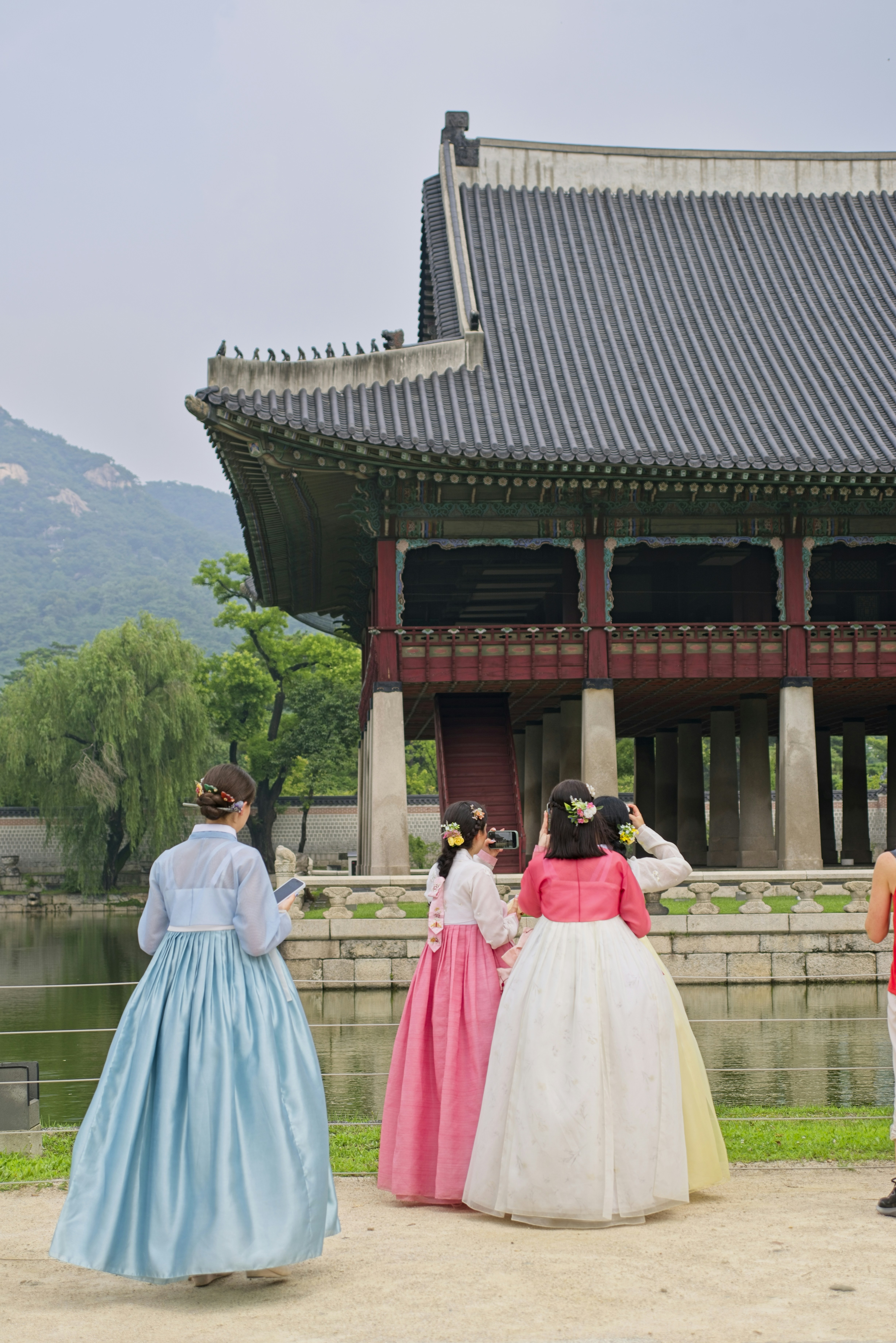 Four women in vibrant hanbok — sky blue, pink, and white — gather at the water's edge before Gyeonghoeru, the palace's grand two-story pavilion raised on stone pillars. Two of them lift smartphones to capture the view, their floral hairpieces and flowing skirts adding color against the muted gray roofline and hazy mountains beyond. Willow trees droop over the still pond in the background. The scene blends candid tourism with picturesque tradition, capturing a relaxed summer afternoon at one of Seoul's most iconic palace landmarks.