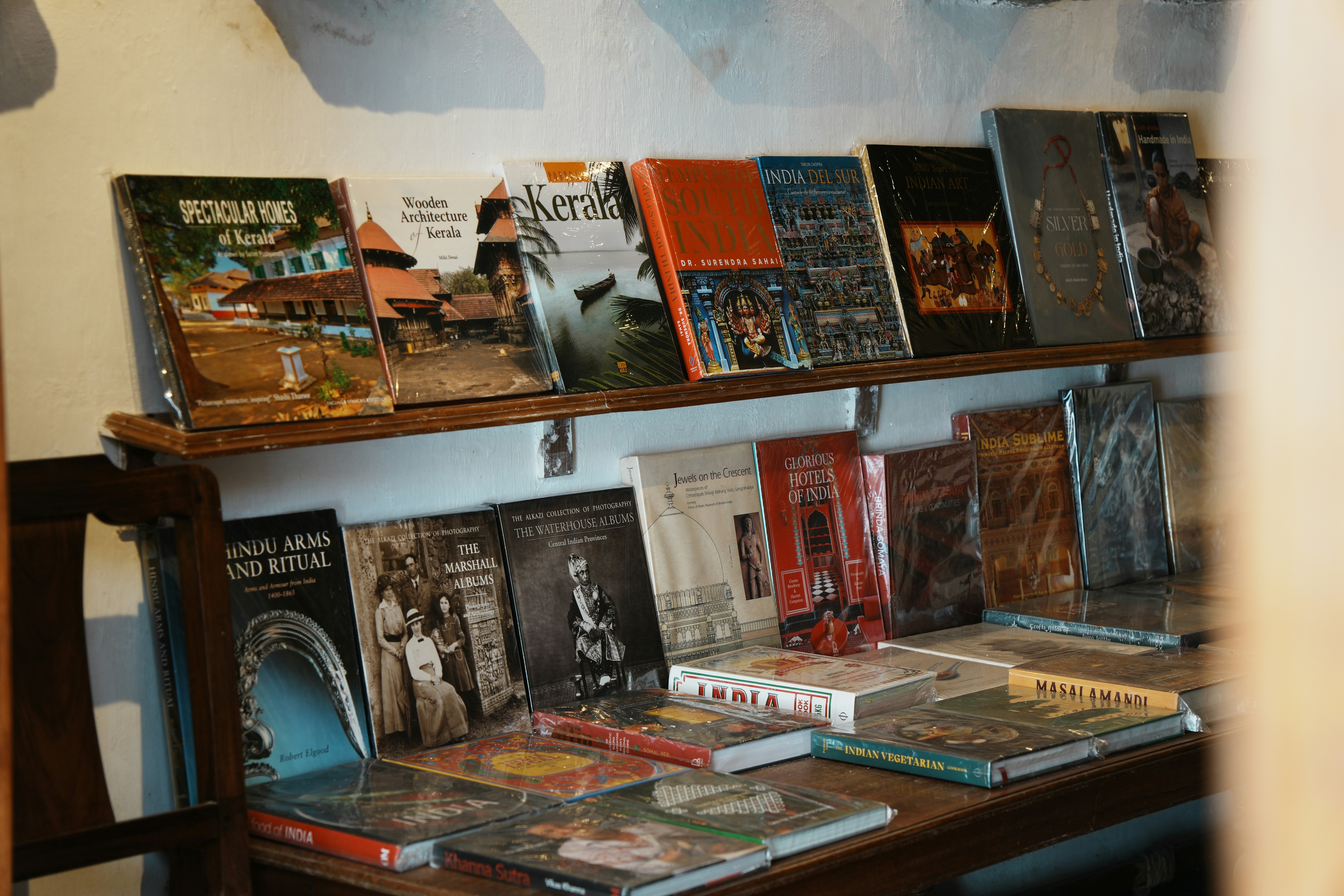 A warm shelf filled with a variety of travel and history books, arranged on two wooden tiers in a softly lit shop.