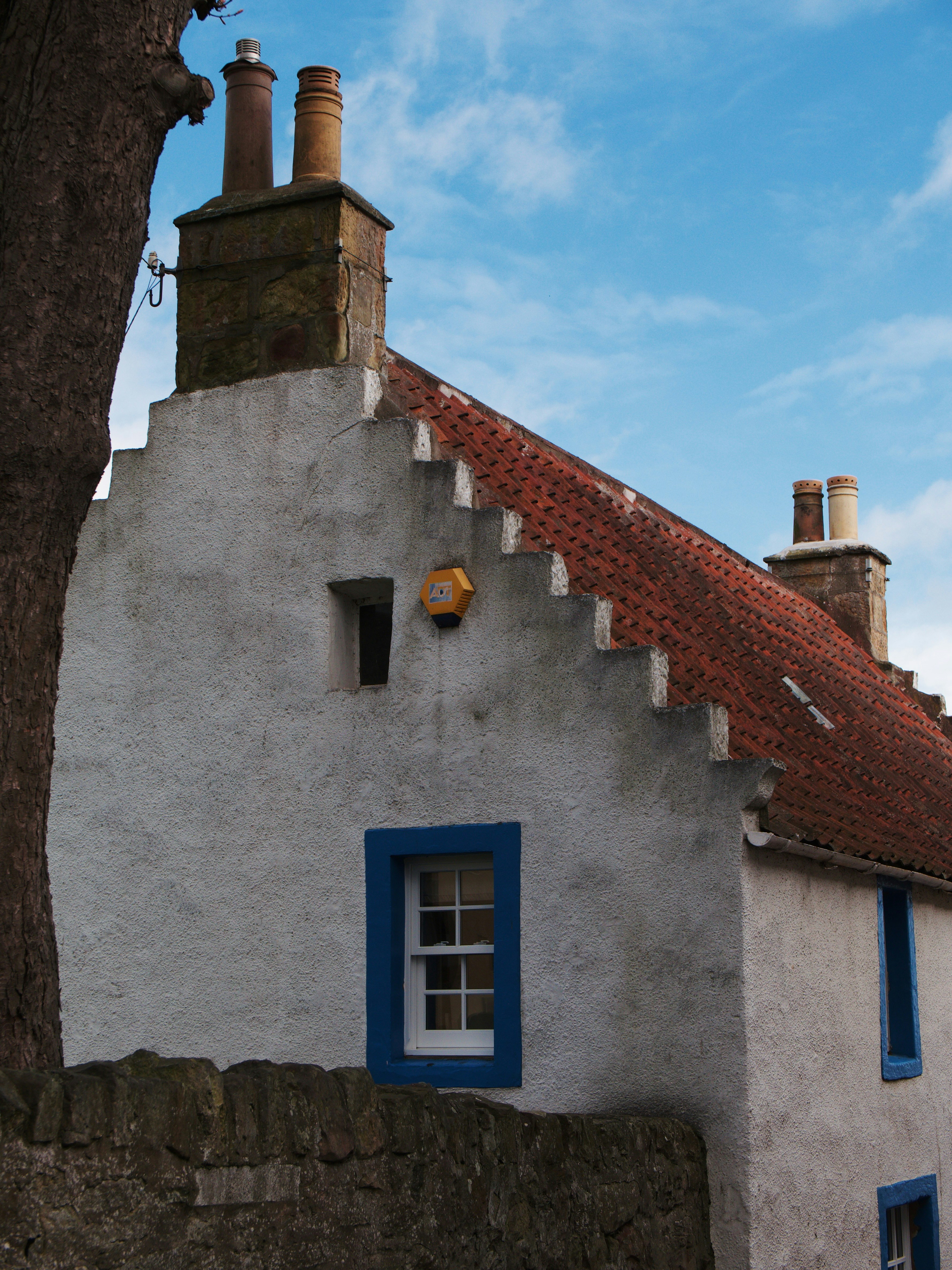 White-washed cottage with a terracotta-tiled roof, blue window frames, and a stepped gable against a bright blue sky.