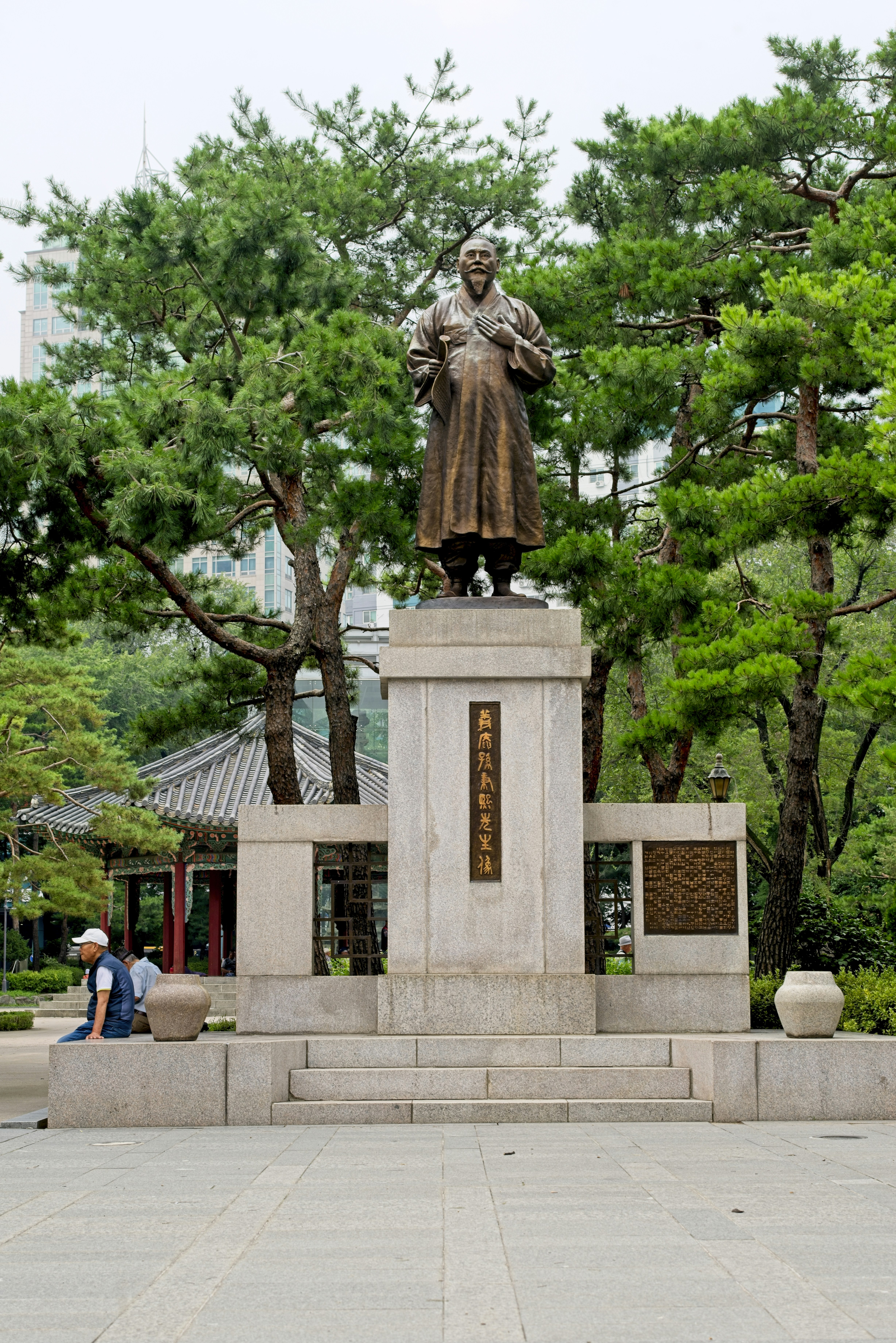 A bronze statue of Son Byeong-hi (의암 손병희), leader of Cheondogyo and a key figure in Korea's March 1st Independence Movement, stands solemnly atop a granite pedestal in Tapgol Park, where the historic 1919 declaration was read. Tall pine trees and a traditional pavilion frame the monument, while modern high-rises rise beyond the greenery. Two elderly men rest in the shade nearby. The peaceful park setting honors a pivotal moment in Korean history amid the quiet bustle of a summer midday in Seoul.