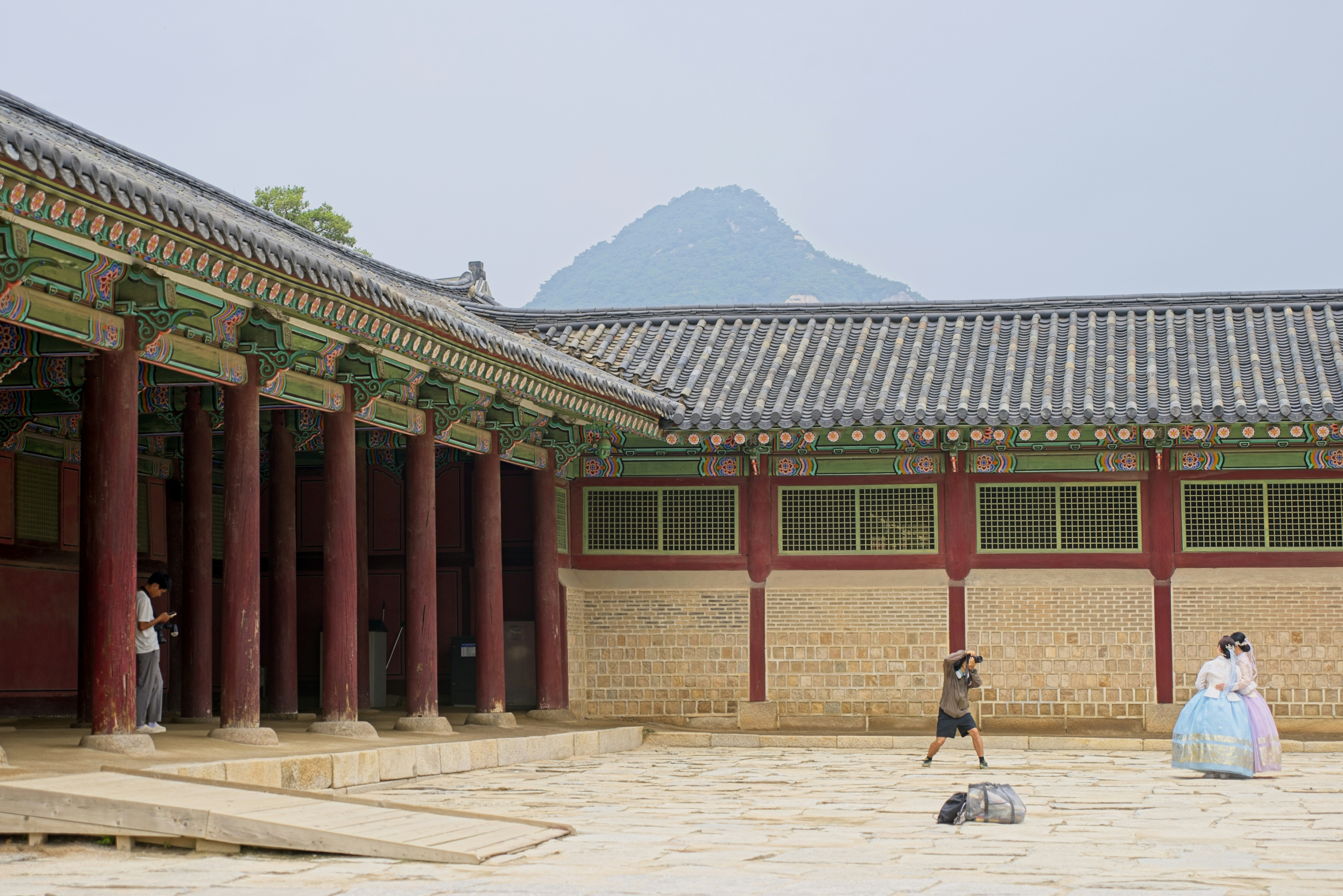 A photographer crouches mid-shot, camera raised toward two women posing in vibrant blue and lavender hanbok against the weathered brick wall of a palace building. Ornately painted eaves and deep red pillars frame the scene, while the jagged peak of Bugaksan rises hazily behind the tiled rooftops. A stone courtyard stretches between the two structures, empty and sunlit under the midday sky. The composition captures a candid behind-the-scenes moment of hanbok photography at Gyeongbokgung Palace, blending tradition, tourism, and architectural grandeur.