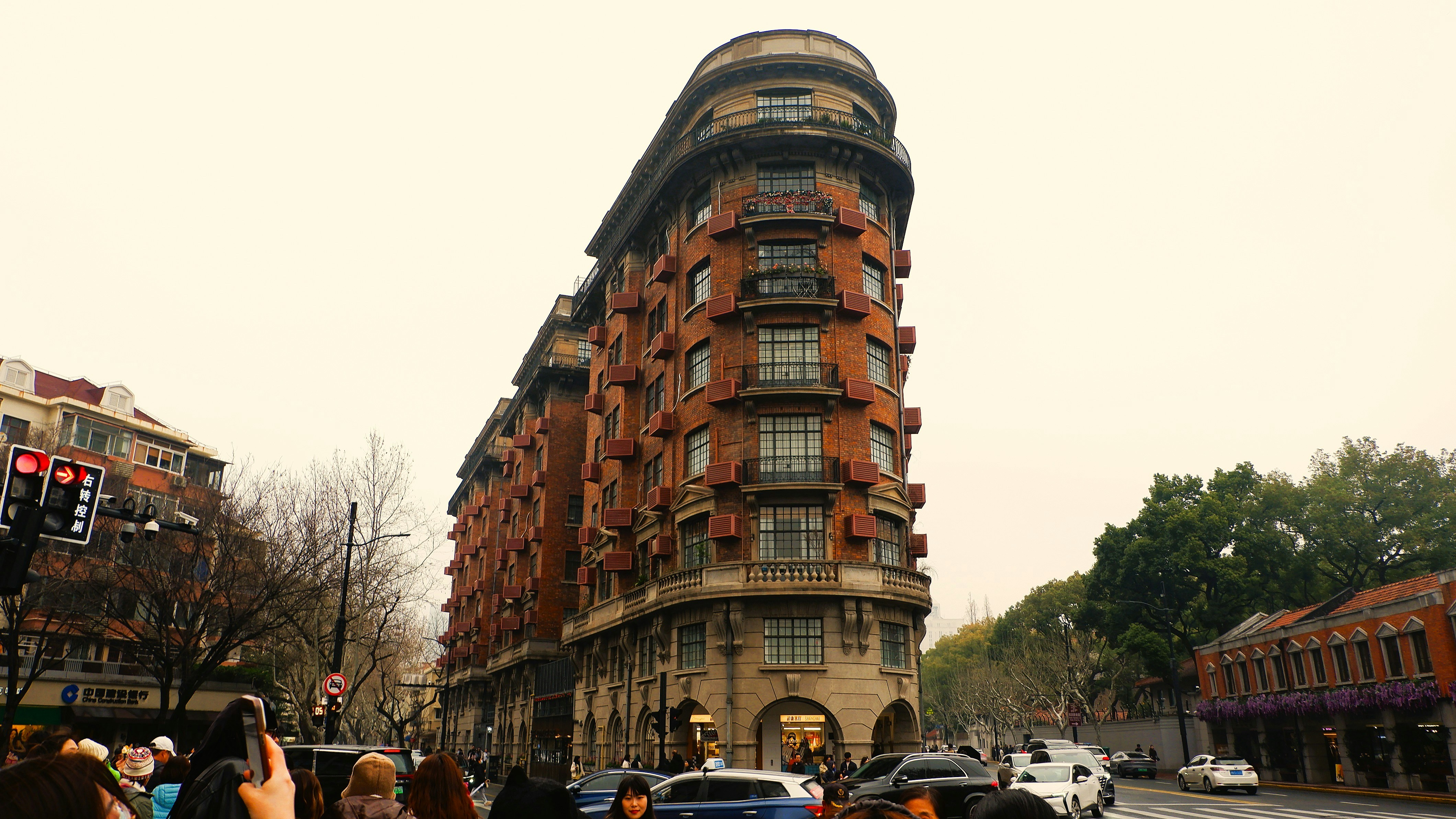 Red-brick curved corner building rising amidst a busy urban scene with pedestrians and cars at its base.