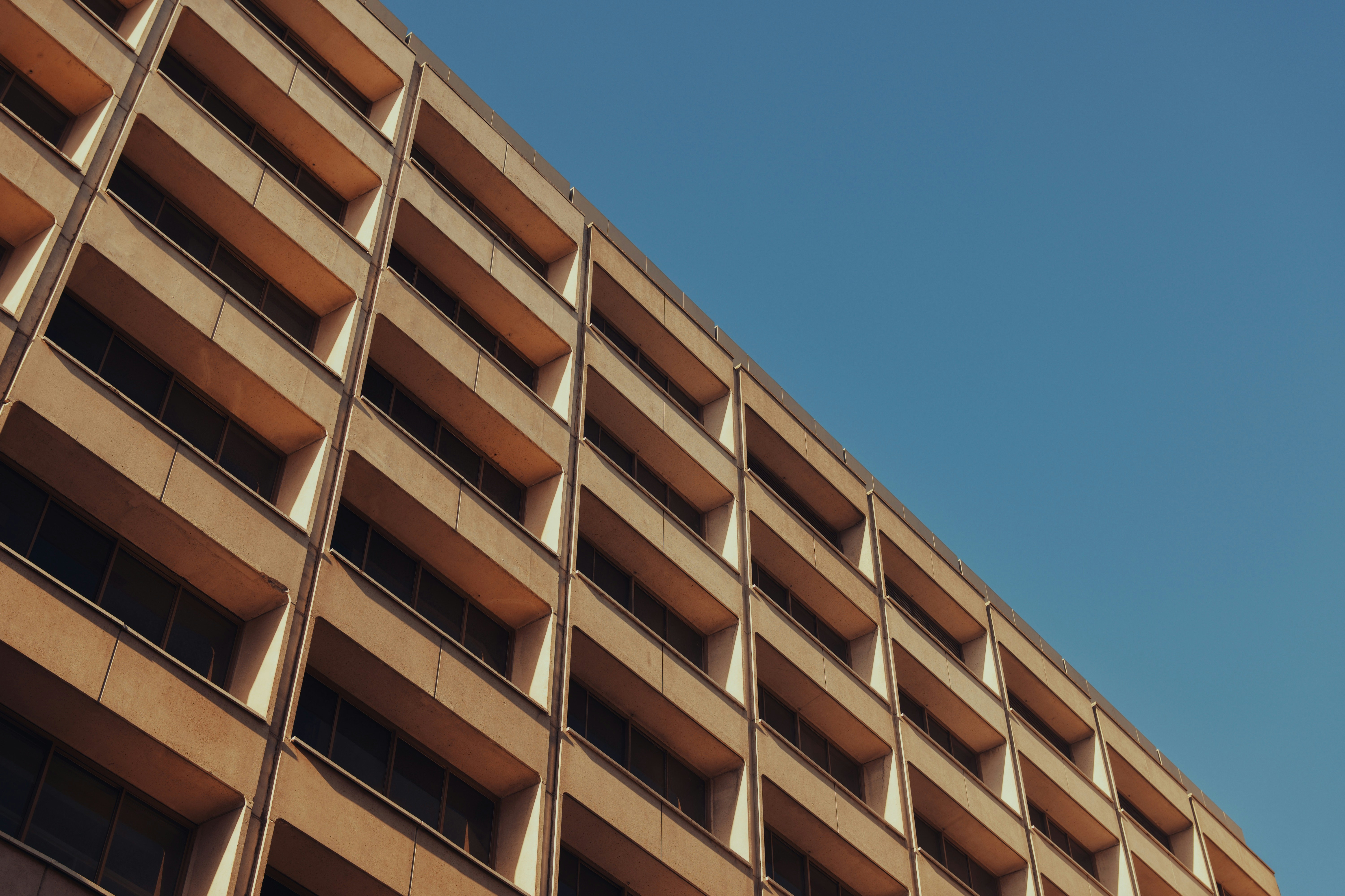 Concrete building facade with repeating balconies set against a clear blue sky; architectural geometry and shadows emphasize a diagonal perspective.