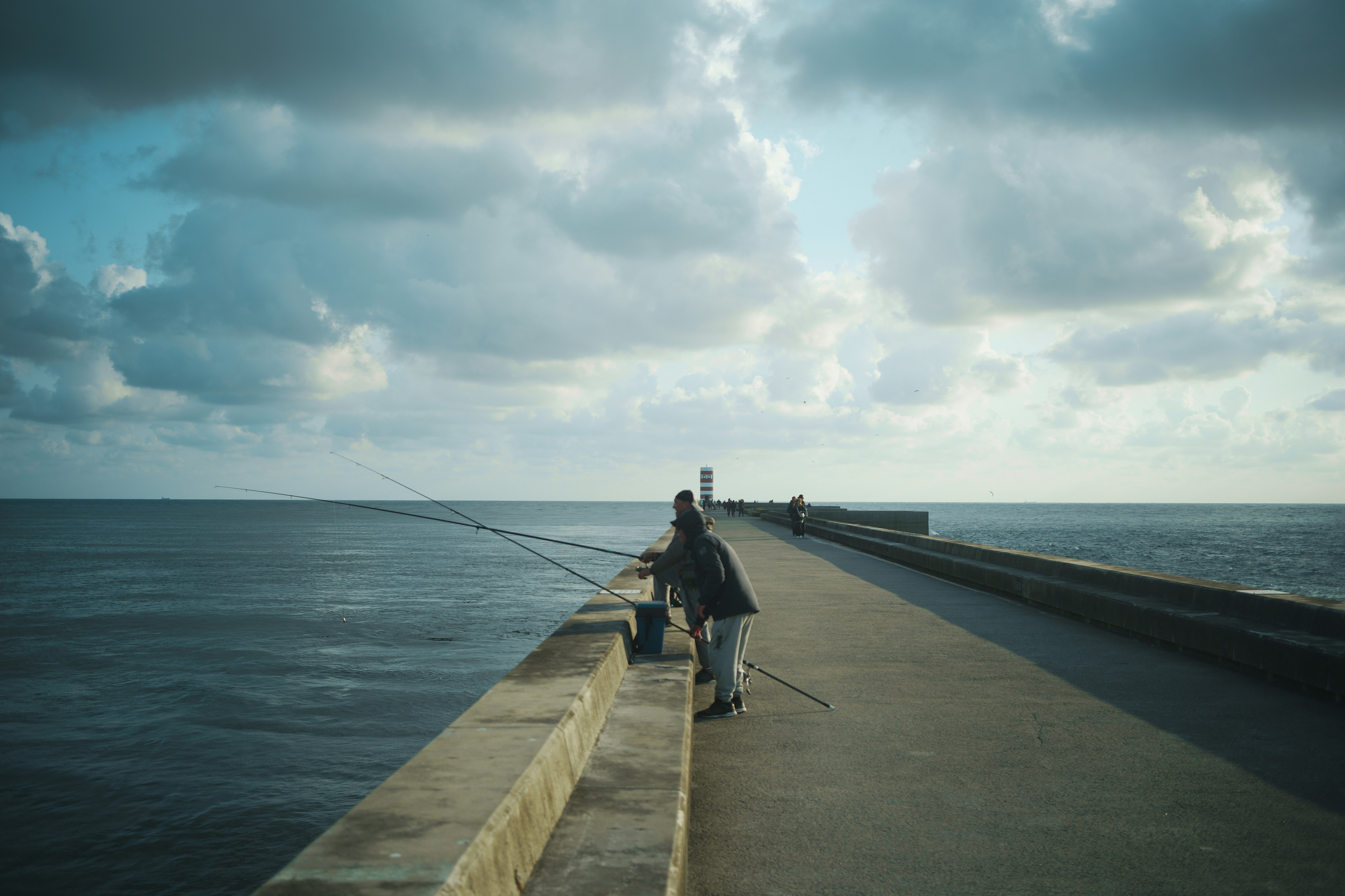 Fisherman leaning on a concrete pier railing extends a fishing rod toward the sea; the scene emphasizes a solitary, contemplative moment along a long pier with moody clouds above.