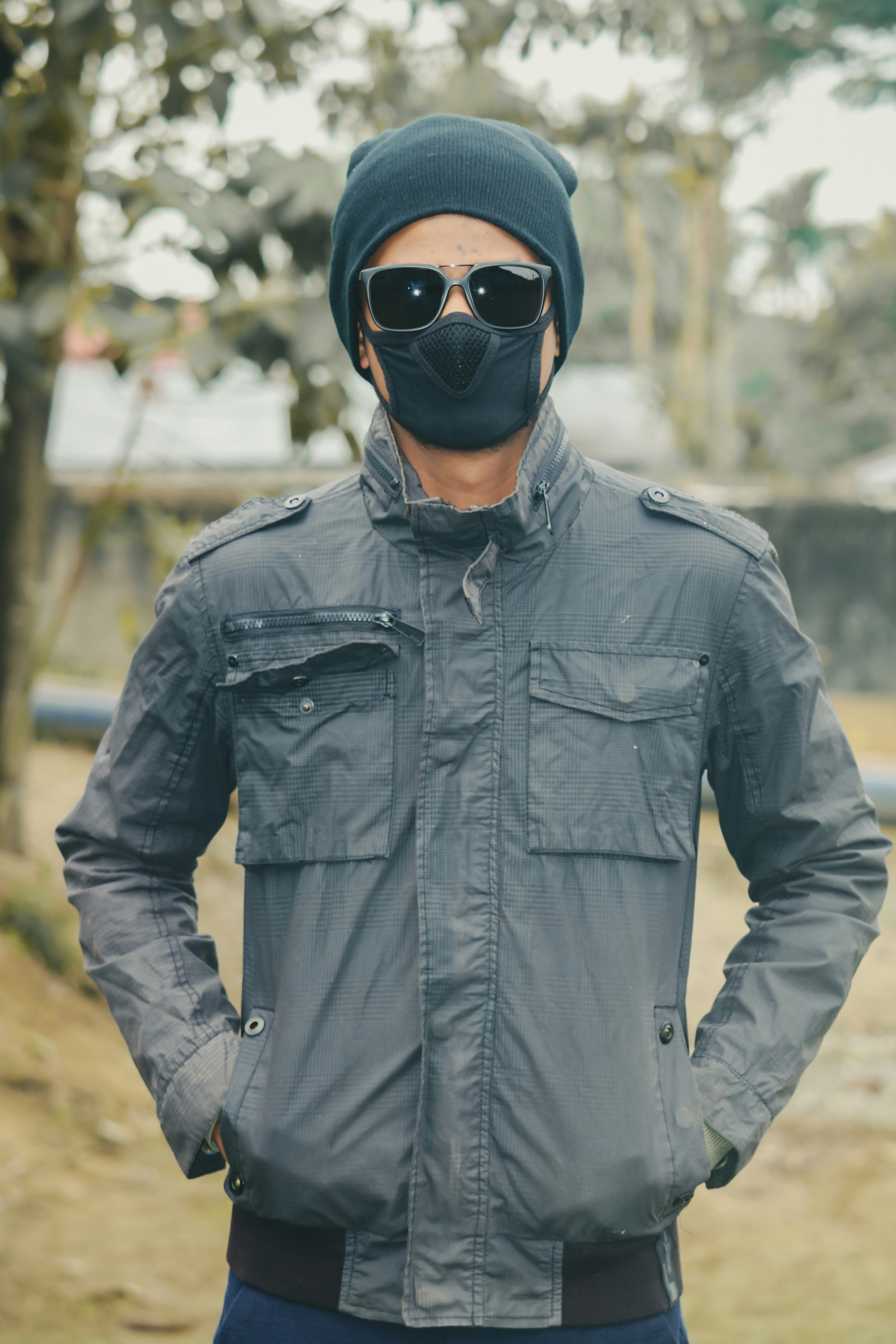 Masked figure wearing dark jacket, beanie, and sunglasses stands outdoors with hands in pockets, conveying a vigilant, urban explorer vibe. (Photograph, portrait-style).