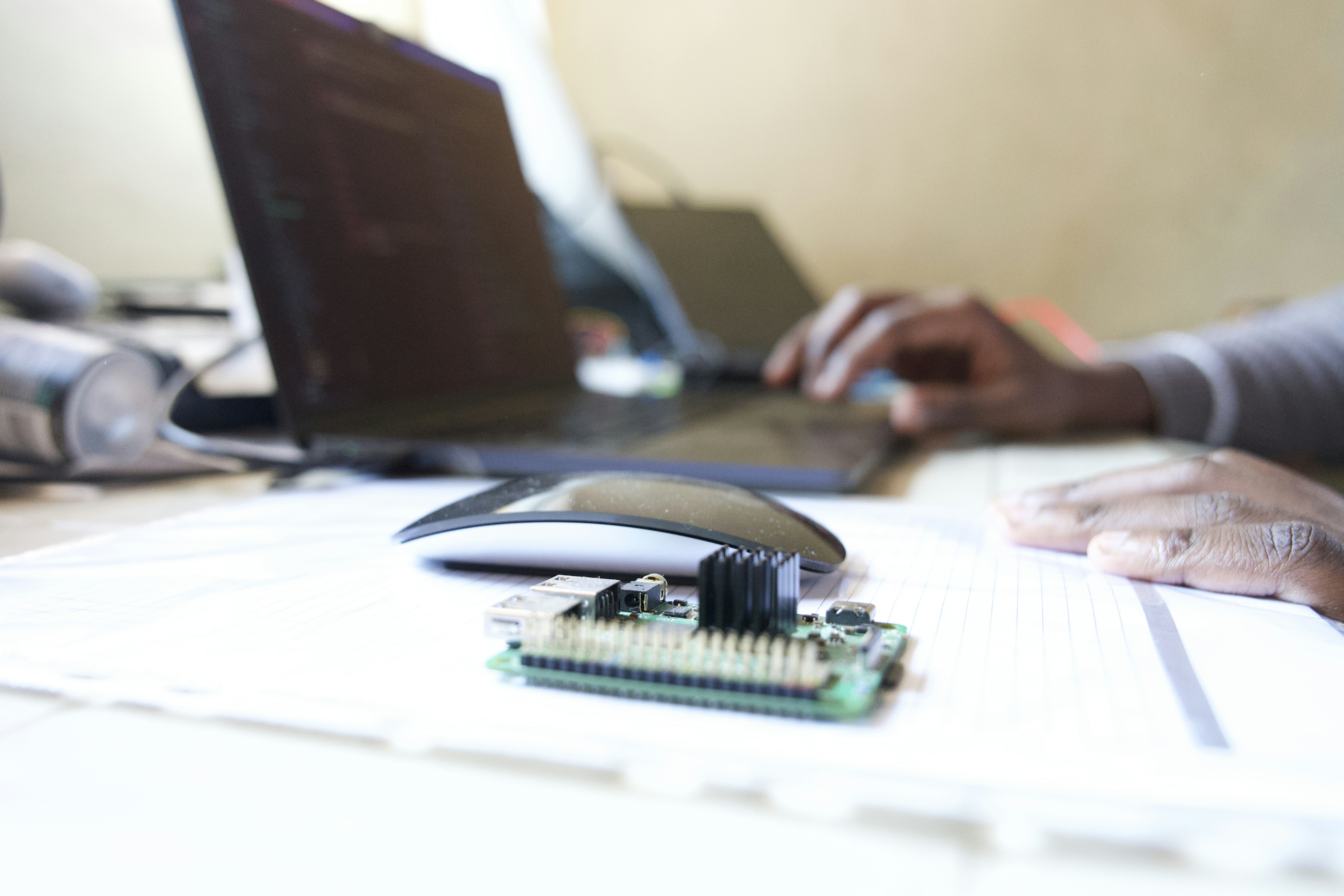 Close-up of a computer mouse, a small circuit board, and a blurred laptop periphery on a desk, conveying a tech workstation scene.