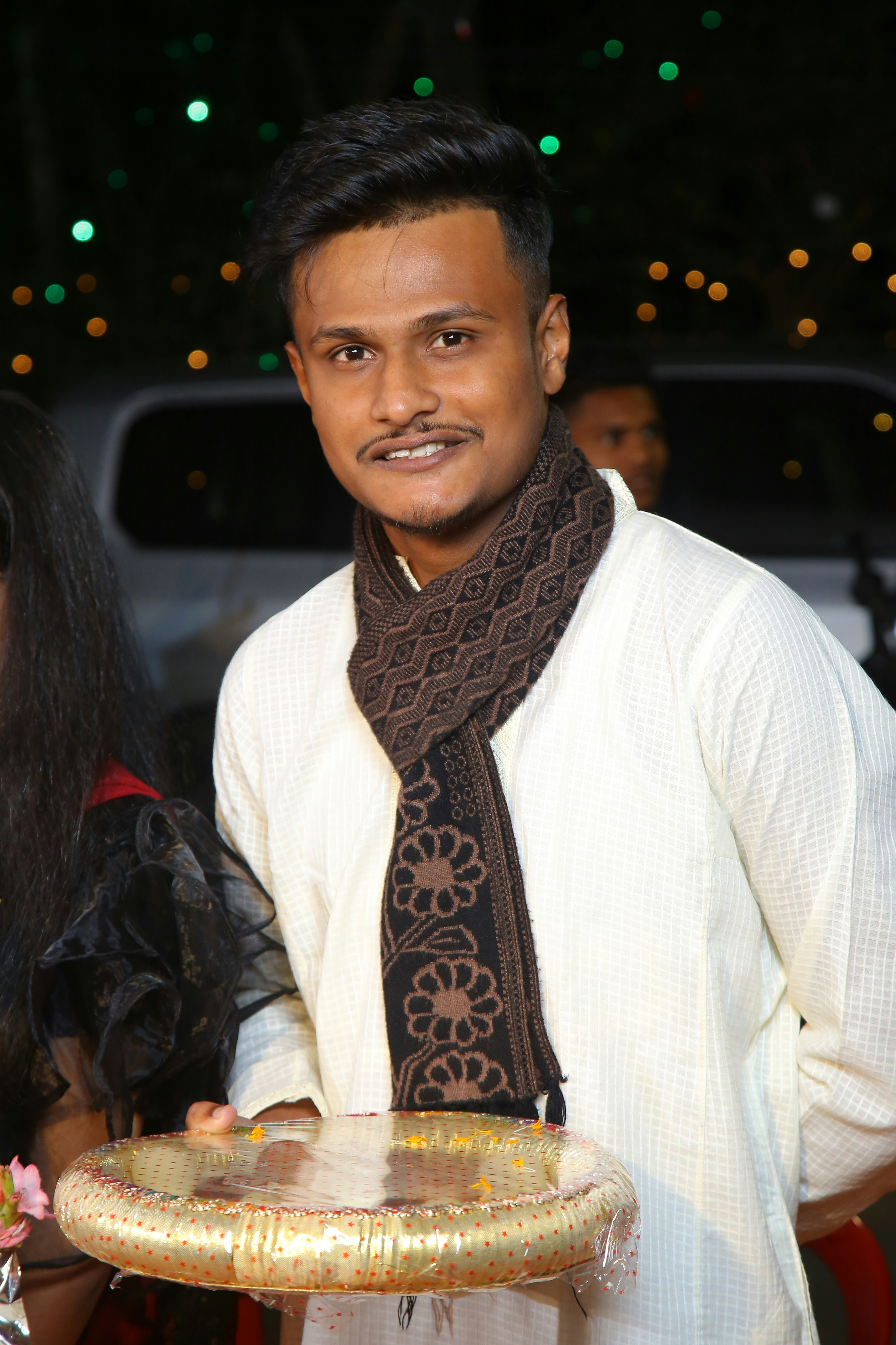 A man smiles toward the camera while presenting a round, decorated plate at a nighttime event; festive lighting creates bokeh in the background.