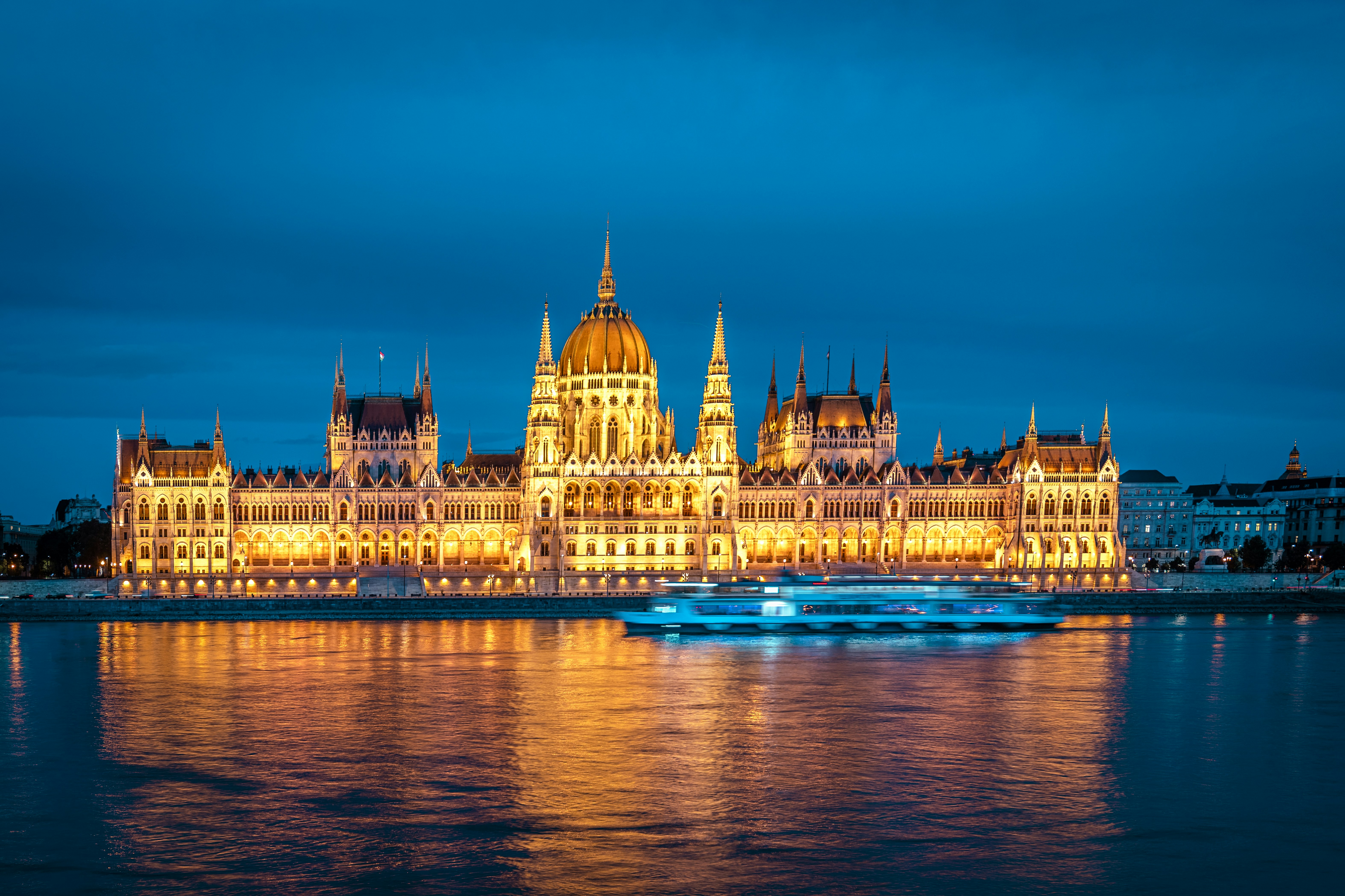 The Hungarian Parliament Building glows along the Danube River at blue hour as a sightseeing boat passes by. A stunning Budapest cityscape featuring historic architecture and waterfront reflections.