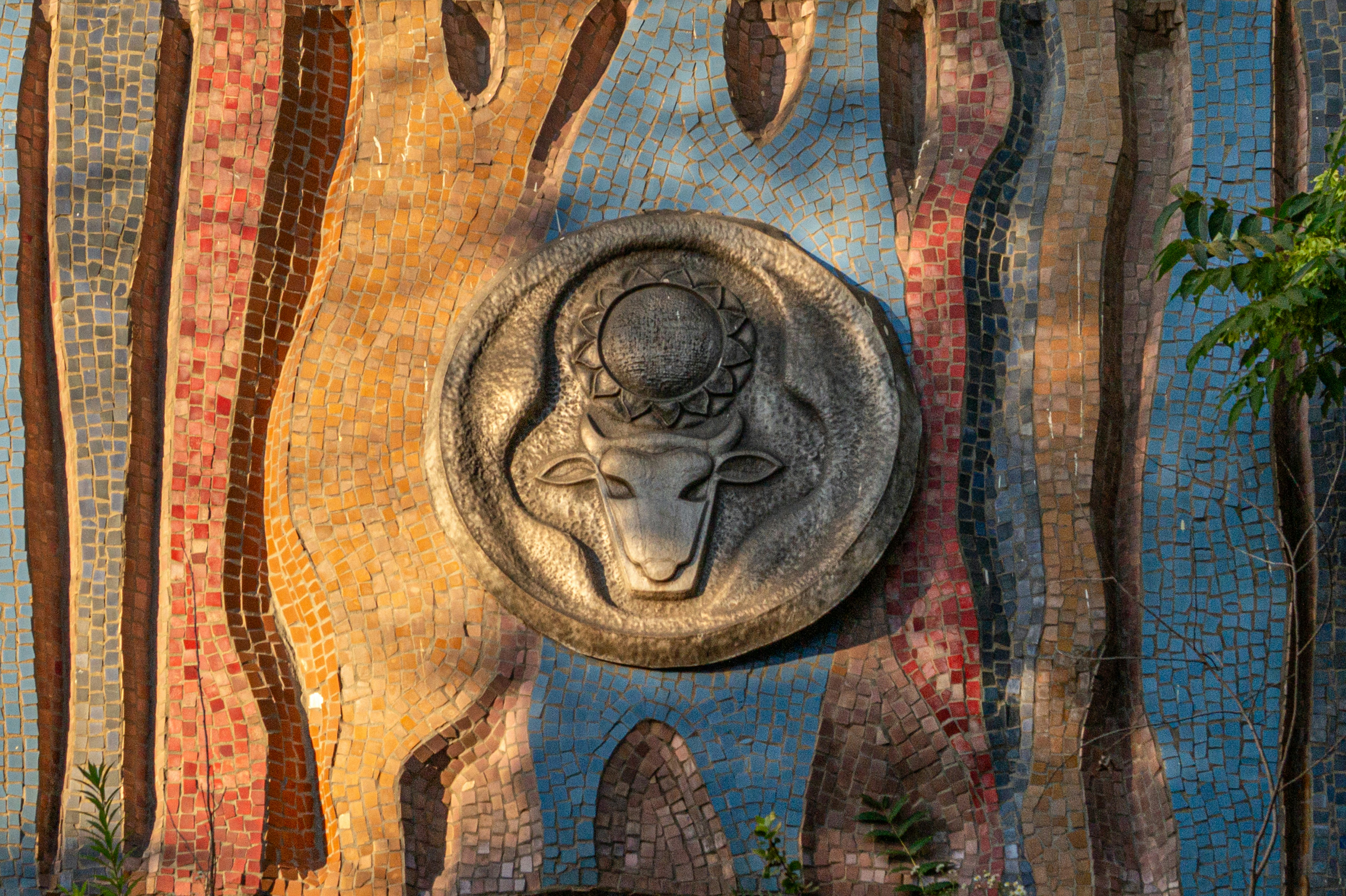 Colorful mosaic wall with a circular ram emblem; sunlit relief sits at the center of wavy ceramic strips, creating a textured focal point. The scene emphasizes architectural art and vibrant tiling.