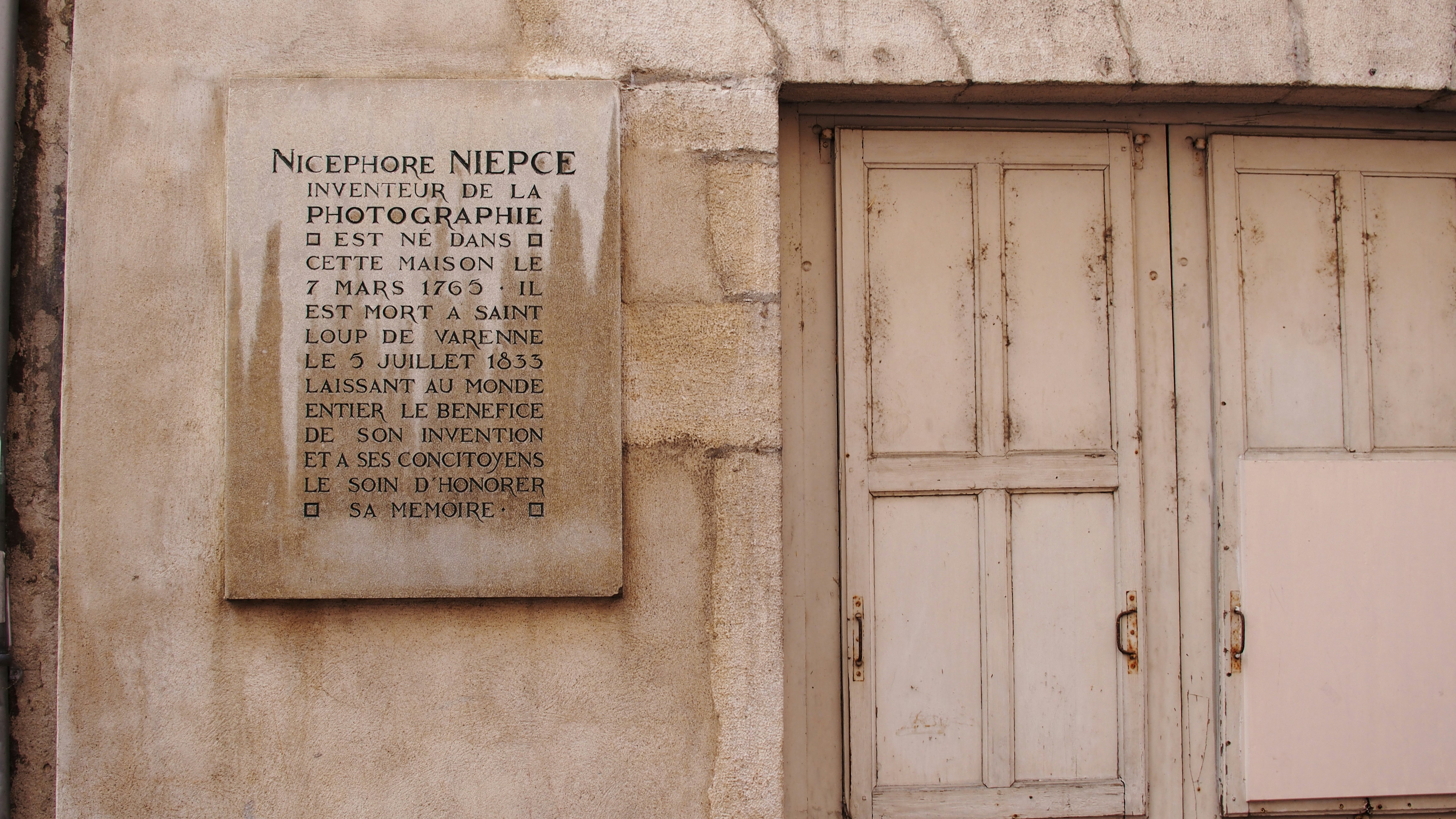 Weathered stone wall with a rectangular plaque beside a worn, wooden door, suggesting a historic or commemorative site. (Logo/inscribed plaque present.)