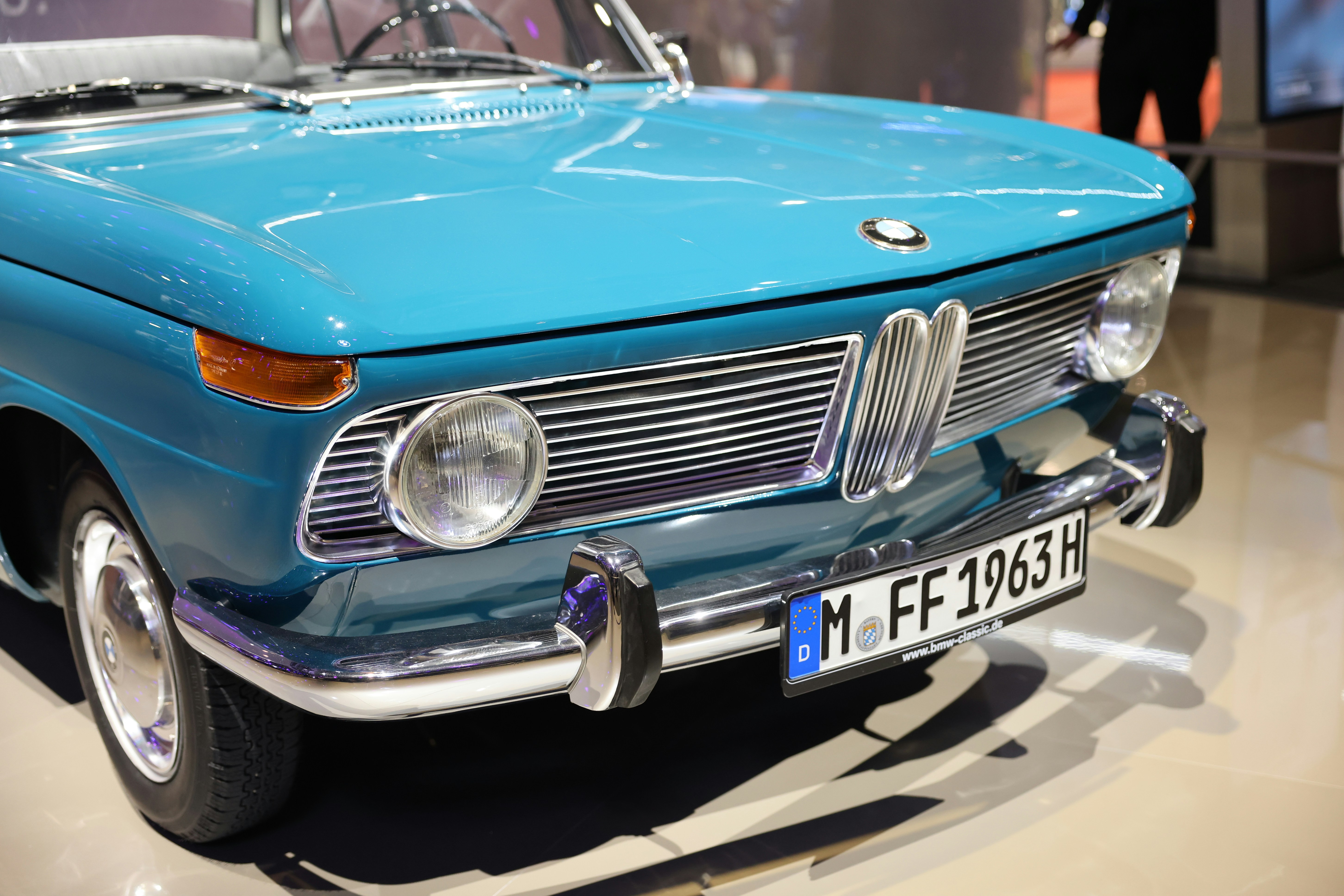 Blue vintage BMW on display with chrome bumper and grille, license plate visible; studio lighting emphasizes polished surfaces.