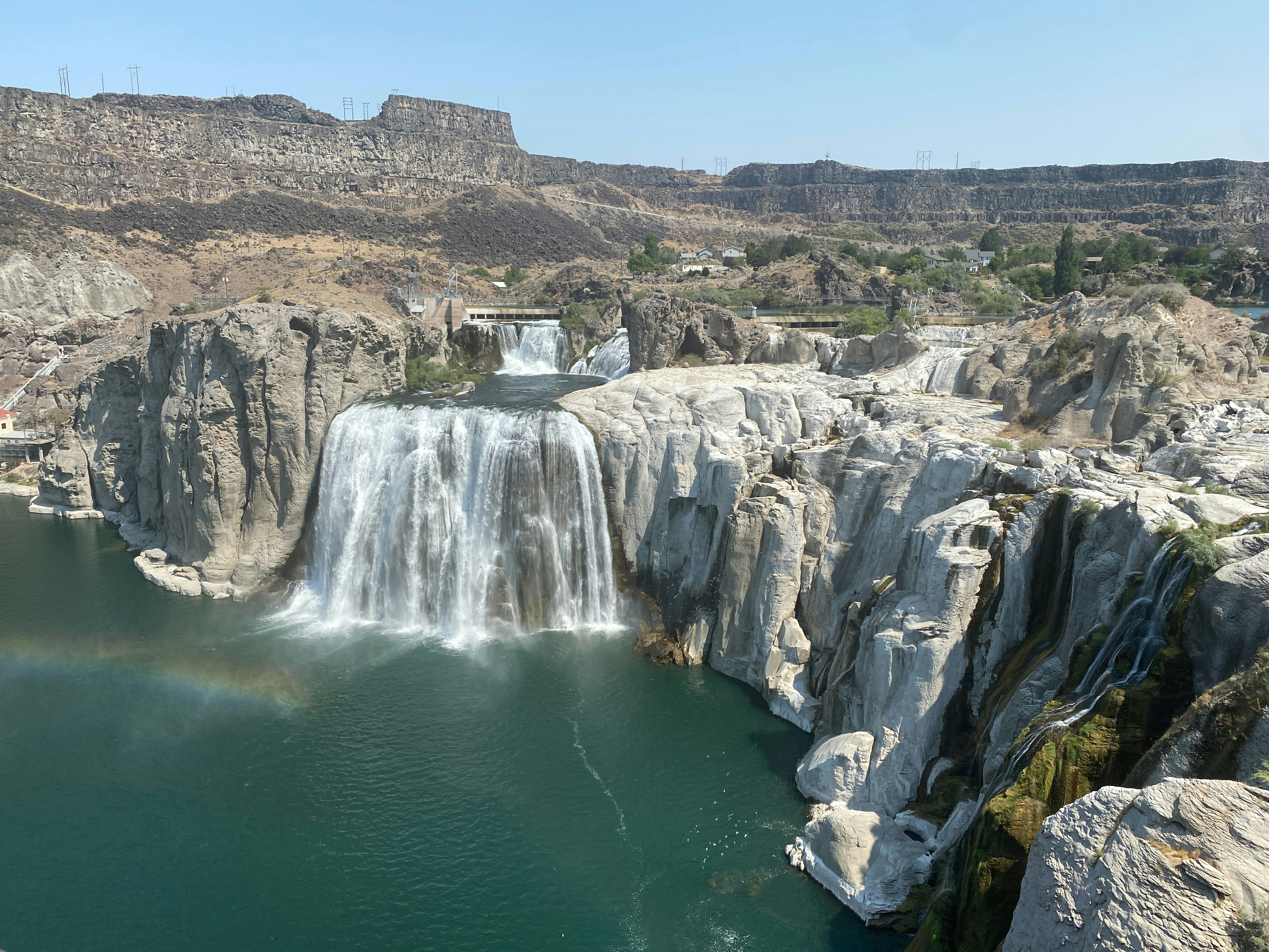 A broad waterfall pours over rugged granite cliffs into a deep turquoise pool, framed by eroded rock faces and sparse vegetation, forming a wide landscape scene.