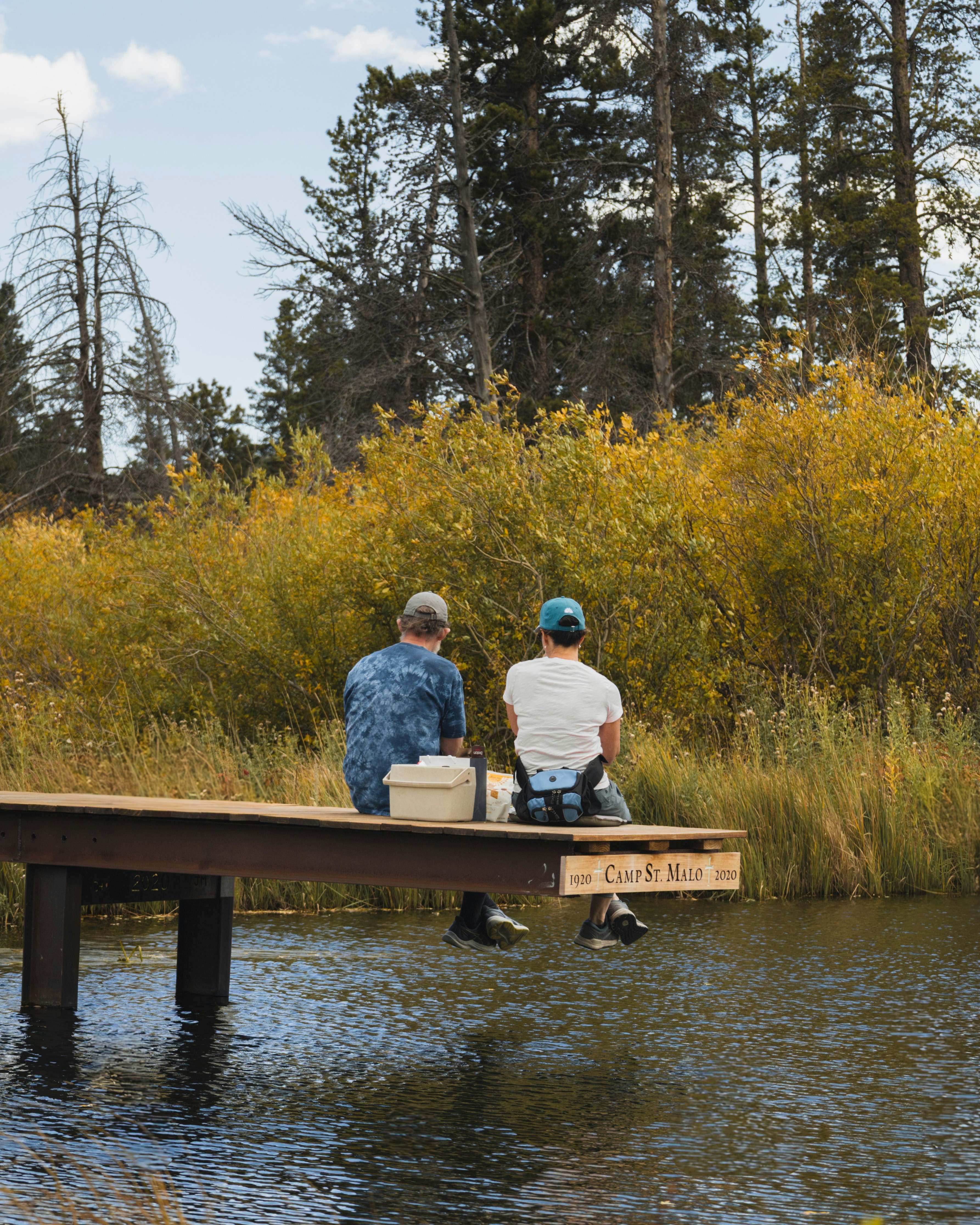 Two anglers on a sunlit dock at the edge of a quiet lake