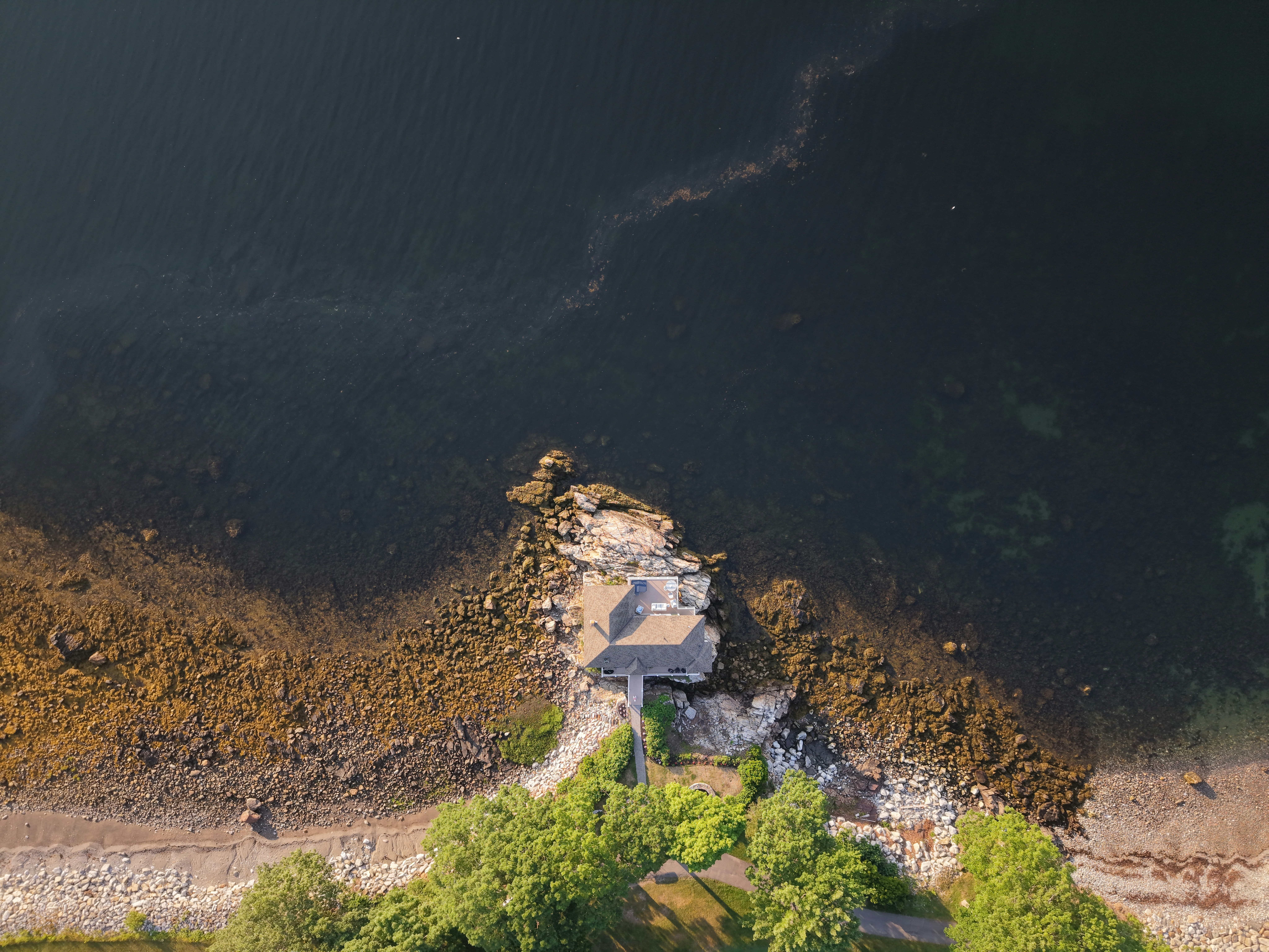Coastal house perched on a rocky jetty with calm water surrounding and a tree-lined shore.