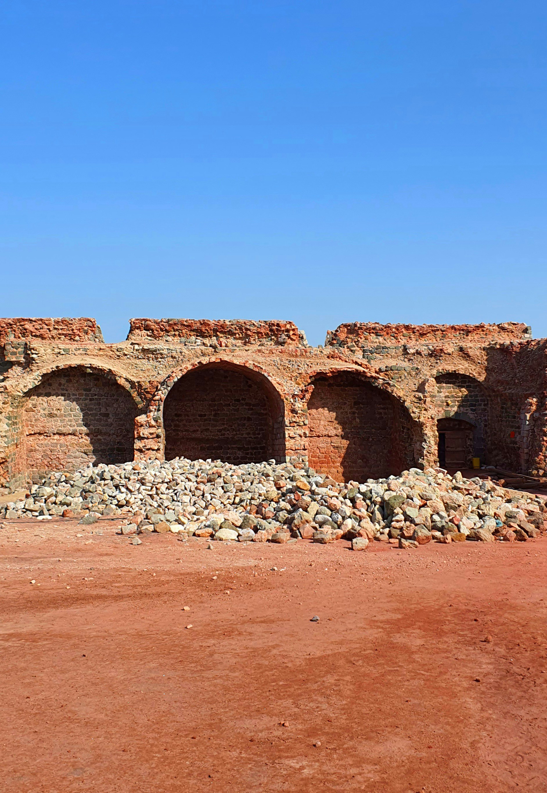 Rough, sun-baked brick arches form a sequence in a desert ruin, with a mound of stones at their base and red soil in the foreground.