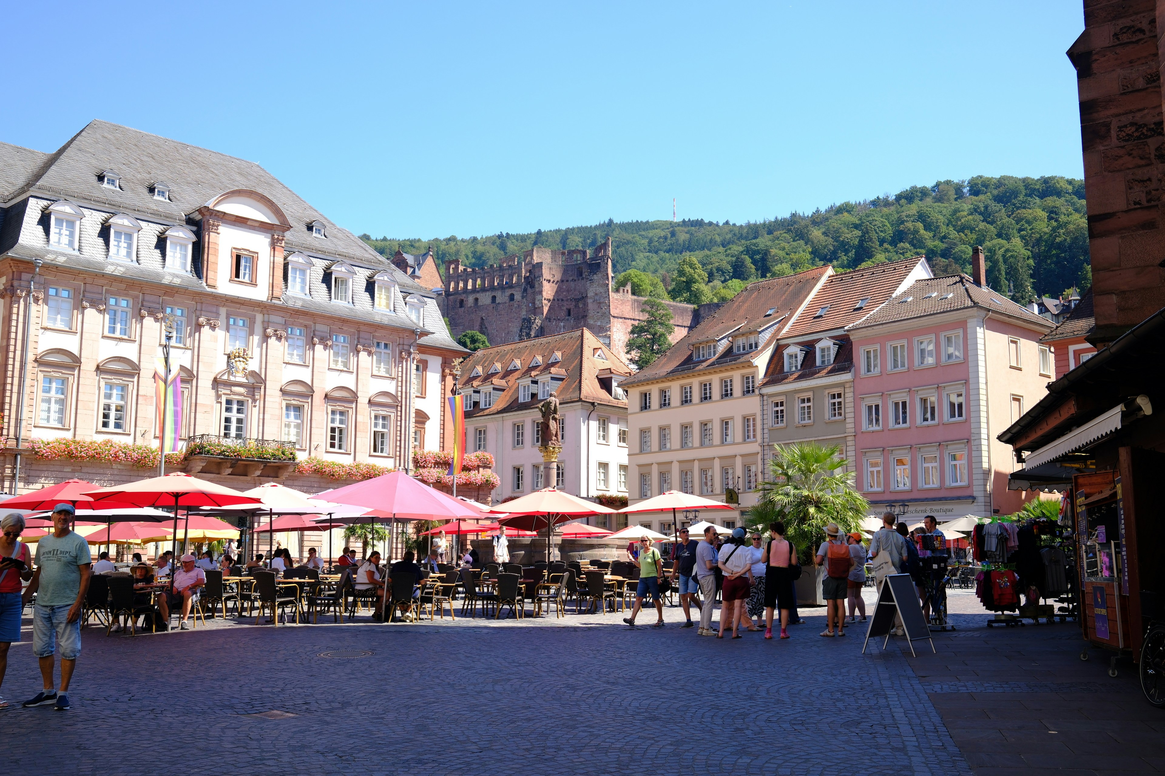 Colorful town square with outdoor cafe seating and pastel façades under a bright blue sky; captures a lively street scene with people gathered near umbrellas.