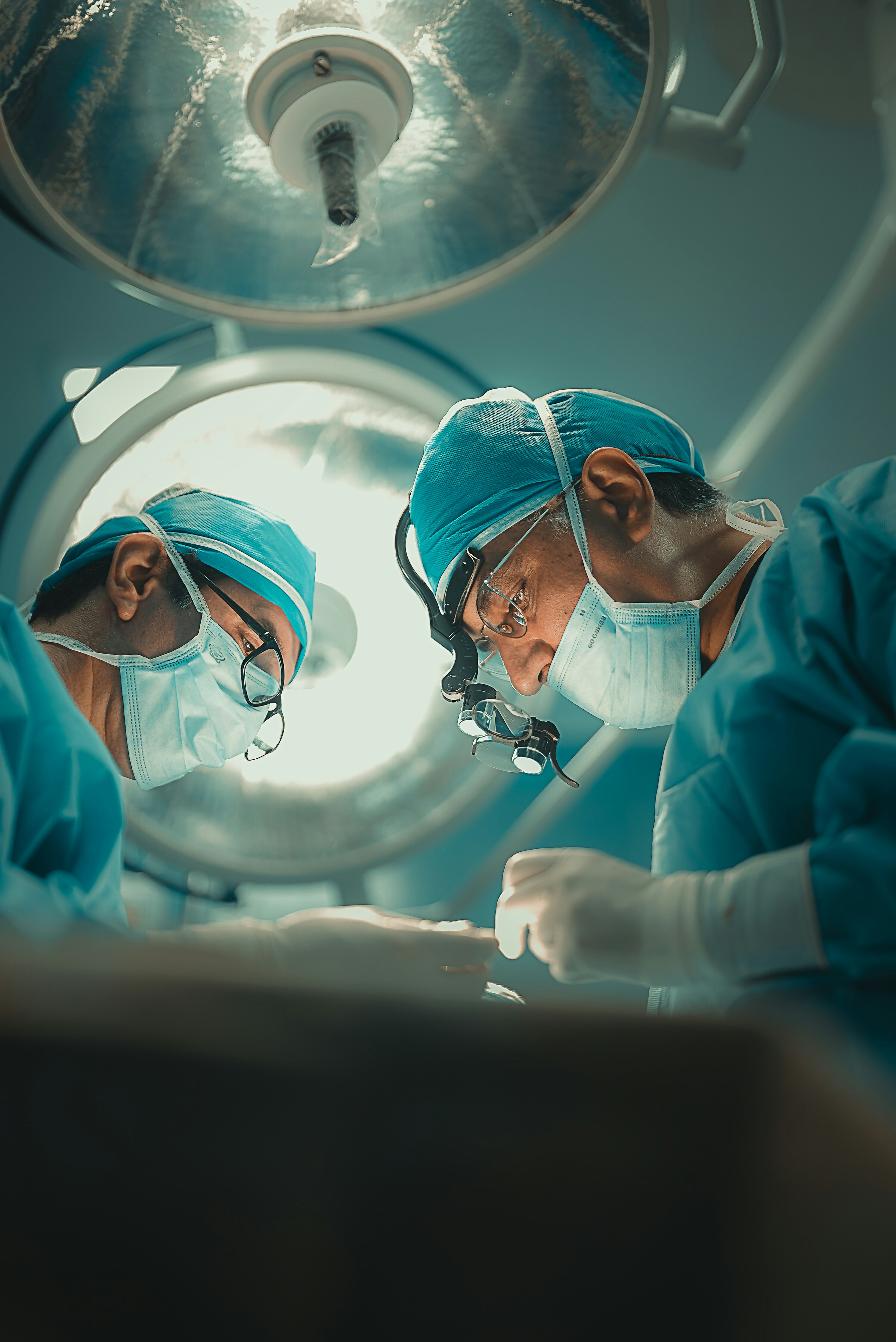 Two surgeons in teal scrubs and masks operate under bright surgical lights, focusing on a procedure with the scene captured from a low angle in the operating room.