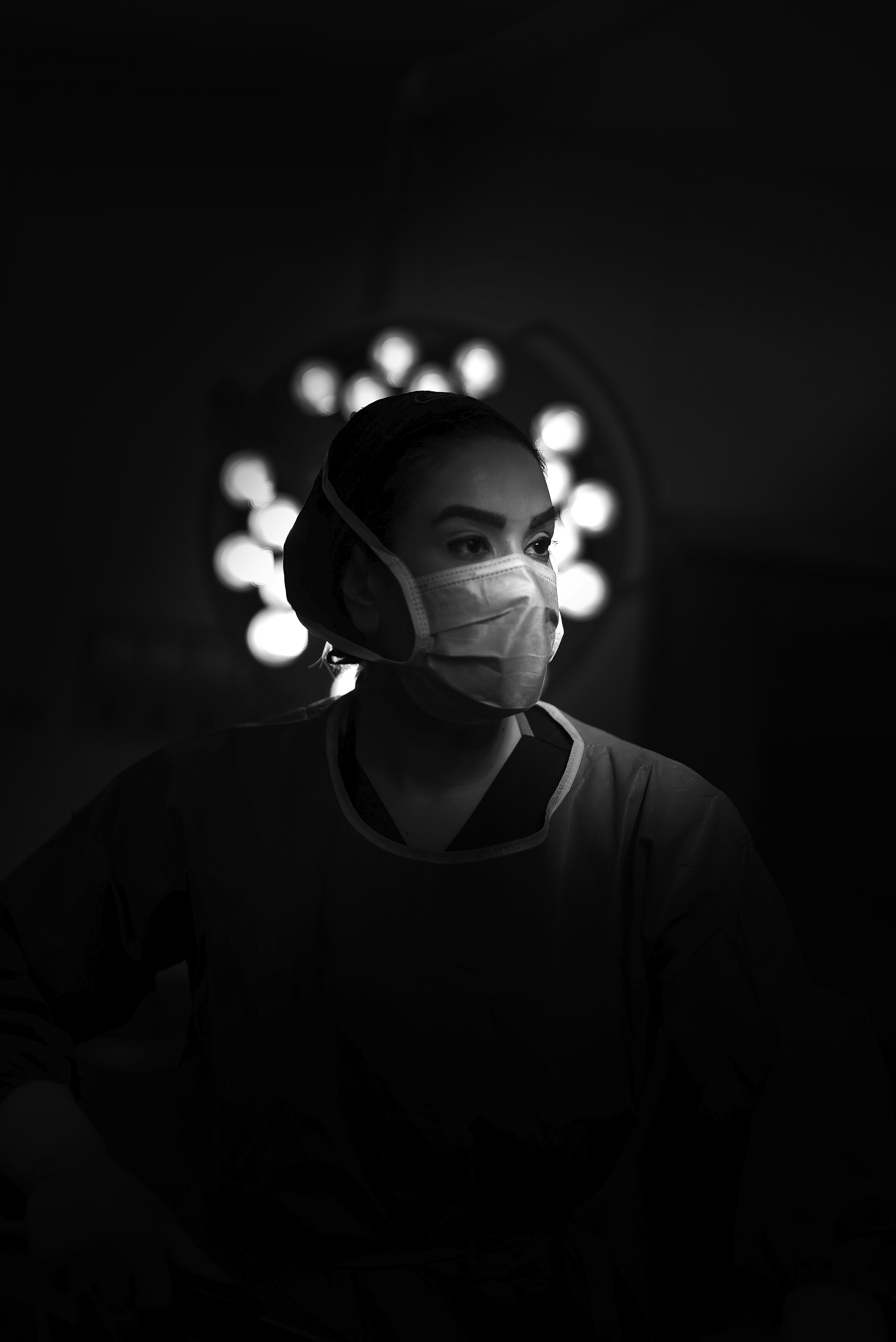 A masked medical professional sits under bright operating room lights, captured in moody black and white with shallow depth showing the face and mask. The image is a photograph.