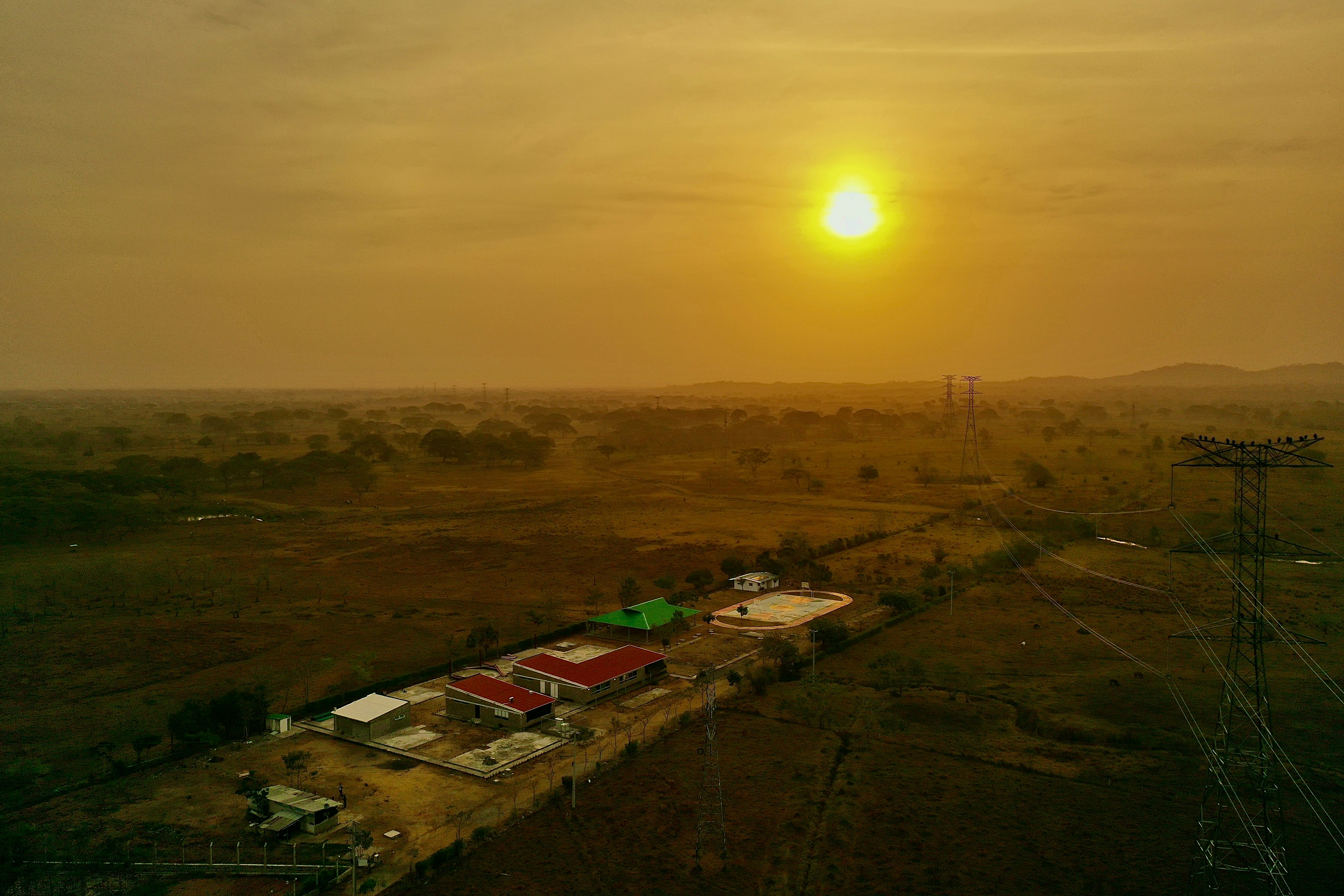 Desolate desert landscape with a small cluster of buildings and radio towers under a warm setting sun. The scene emphasizes aerial perspective and remote infrastructure.