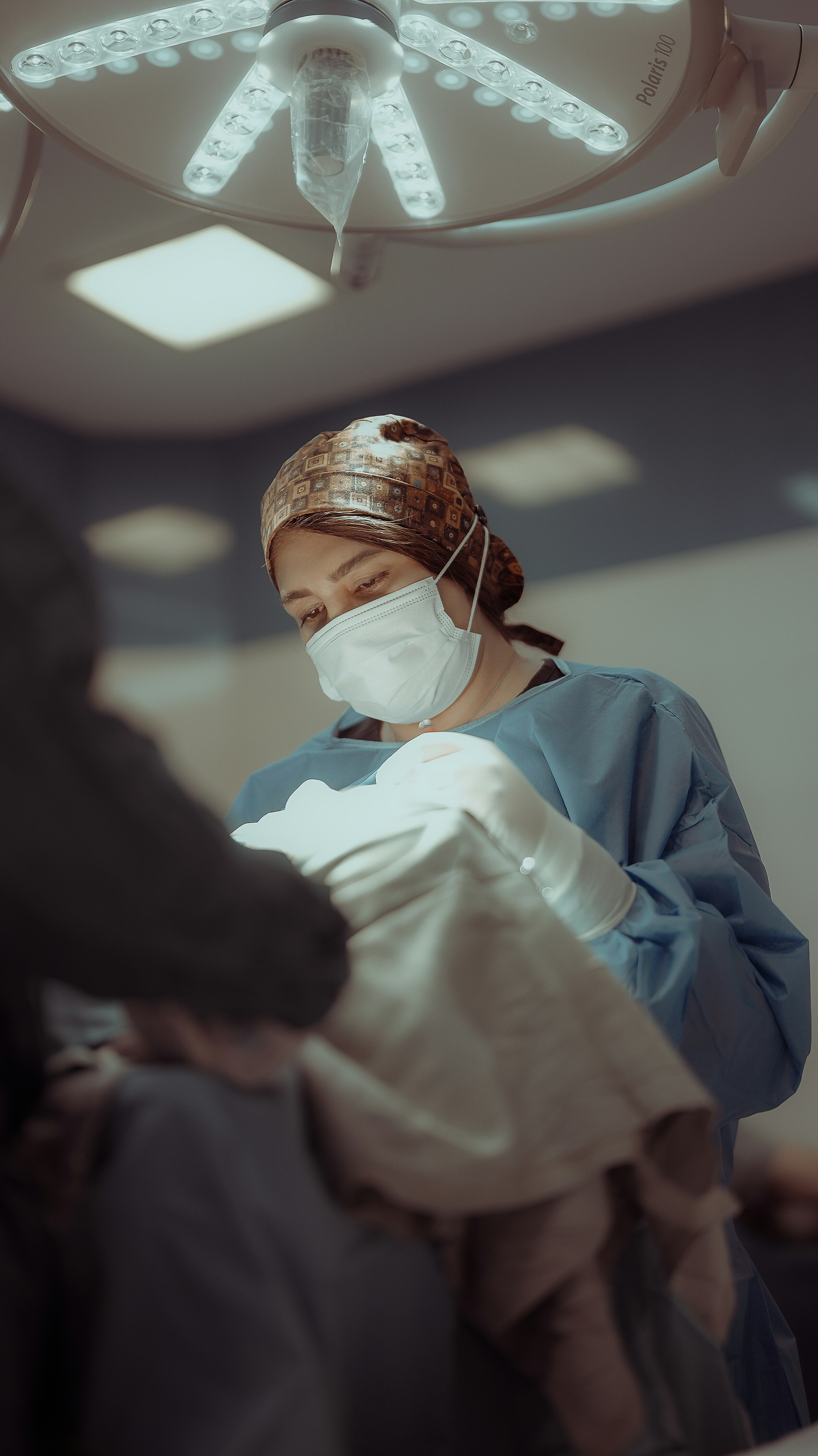 A masked surgeon in blue scrubs and a cap under a surgical lamp prepares to perform, with gloved hands and draped fabric in the foreground.