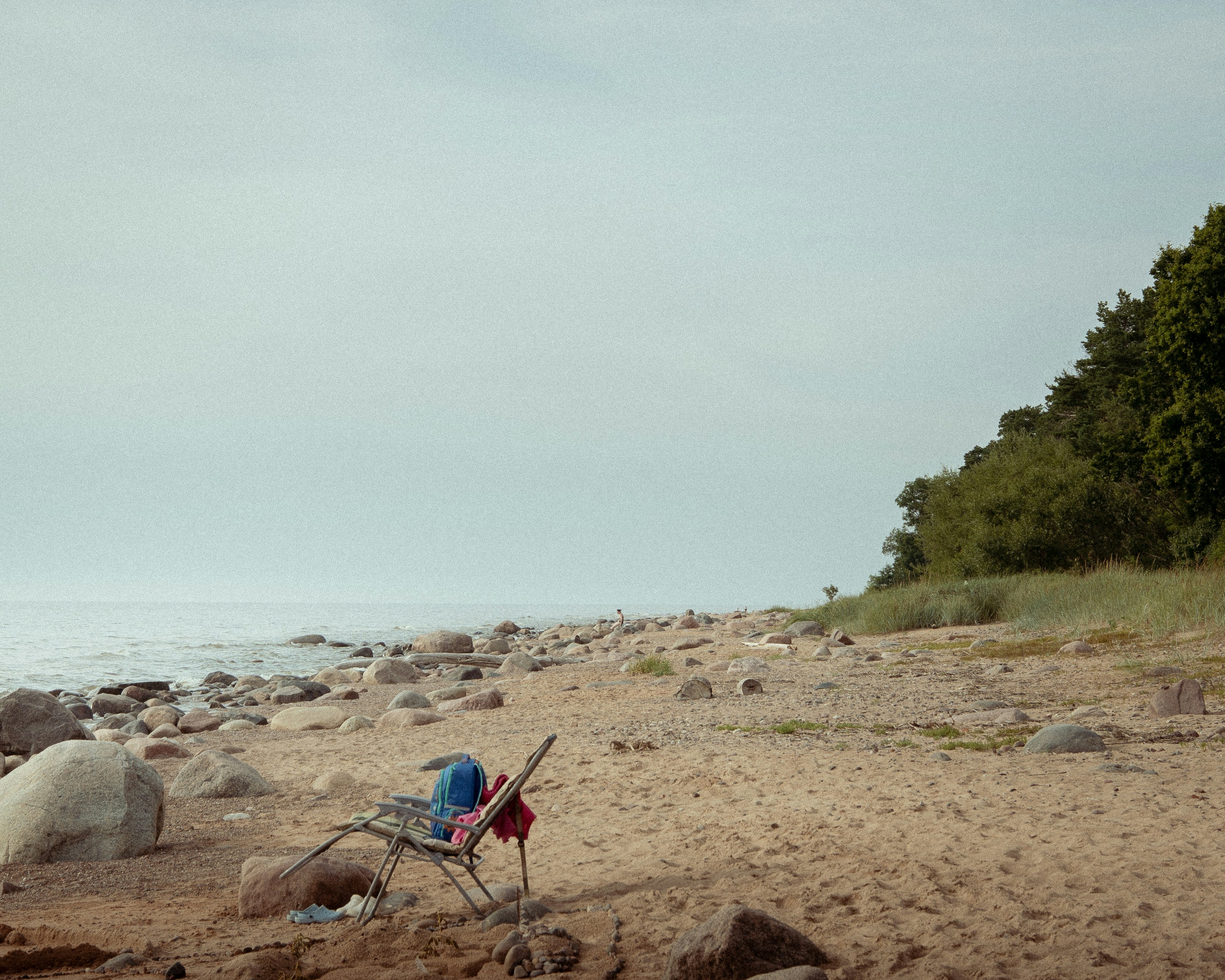 Sandy beach with scattered rocks, a lone folding chair, and a blue bag near the waterline flanked by a grassy dune and trees in the distance. A calm, open shoreline scene.