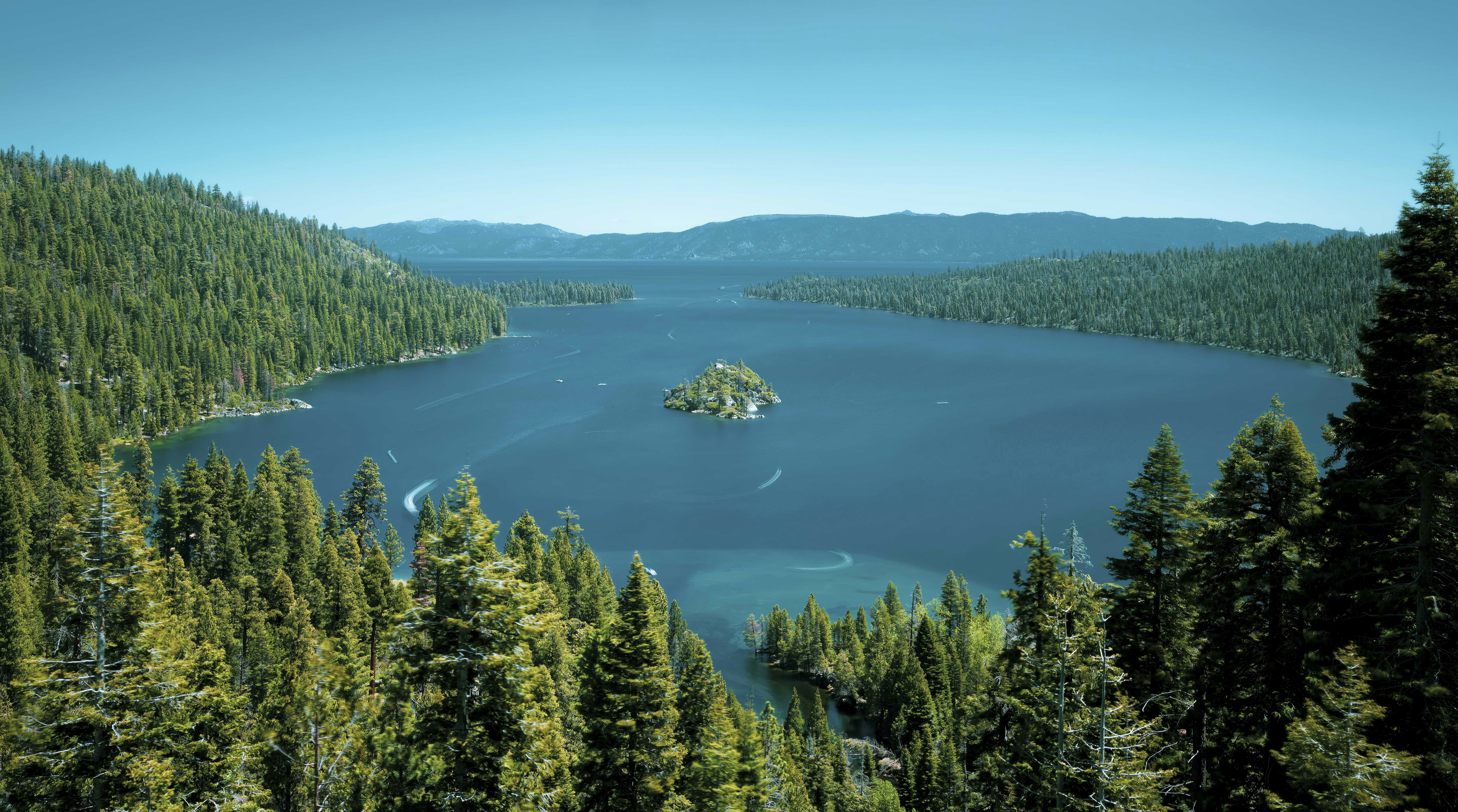 Azure lake waters cradle a small island surrounded by conifer-clad cliffs, framed by a pine-carved canyon under a bright sky.