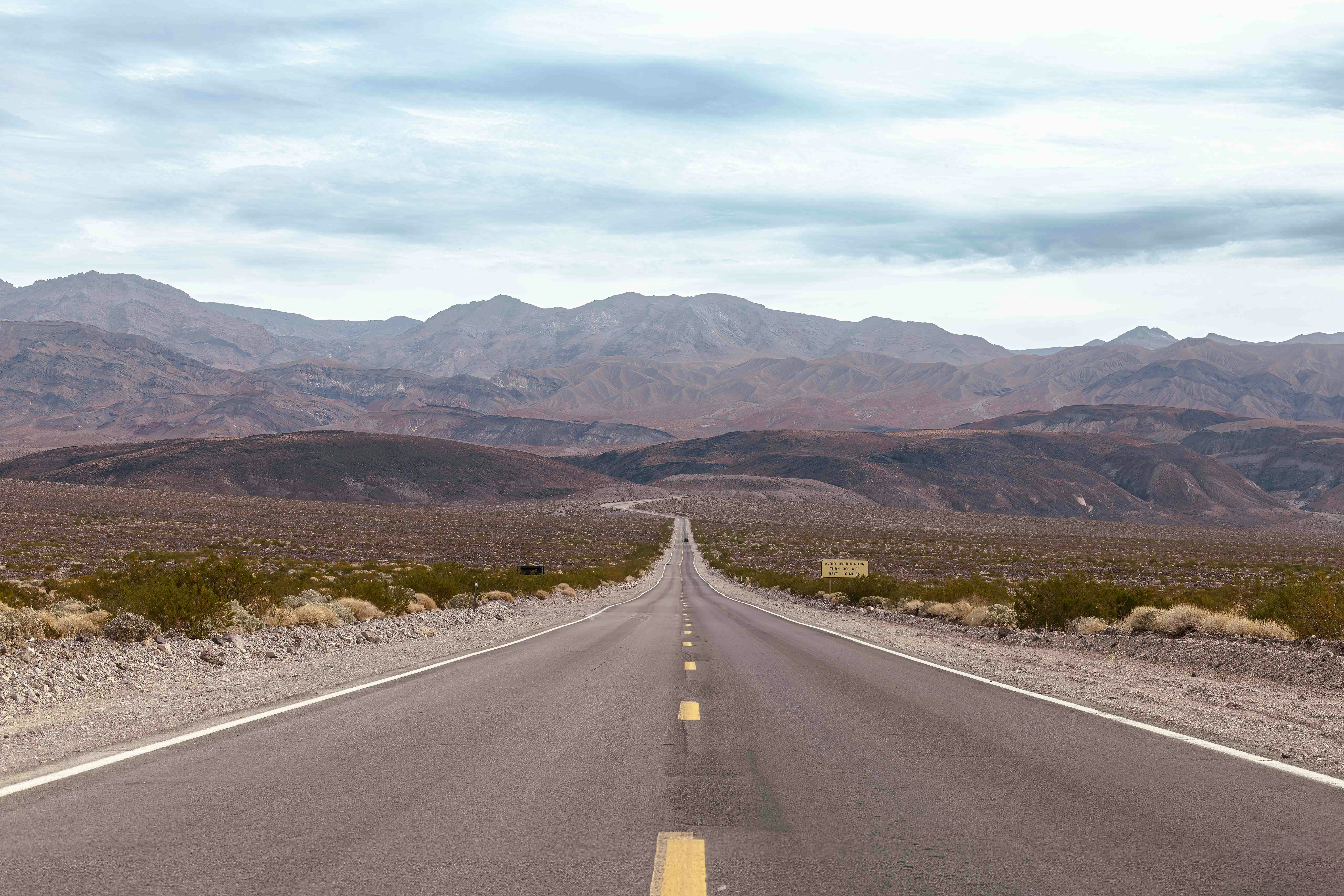 A straight desert highway stretches across Death Valley National Park toward the rugged Panamint Range, creating a striking composition of leading lines, expansive desert terrain, and dramatic mountain scenery beneath a cloud-filled sky. This iconic American Southwest landscape captures the solitude and scale of one of the hottest places on Earth.