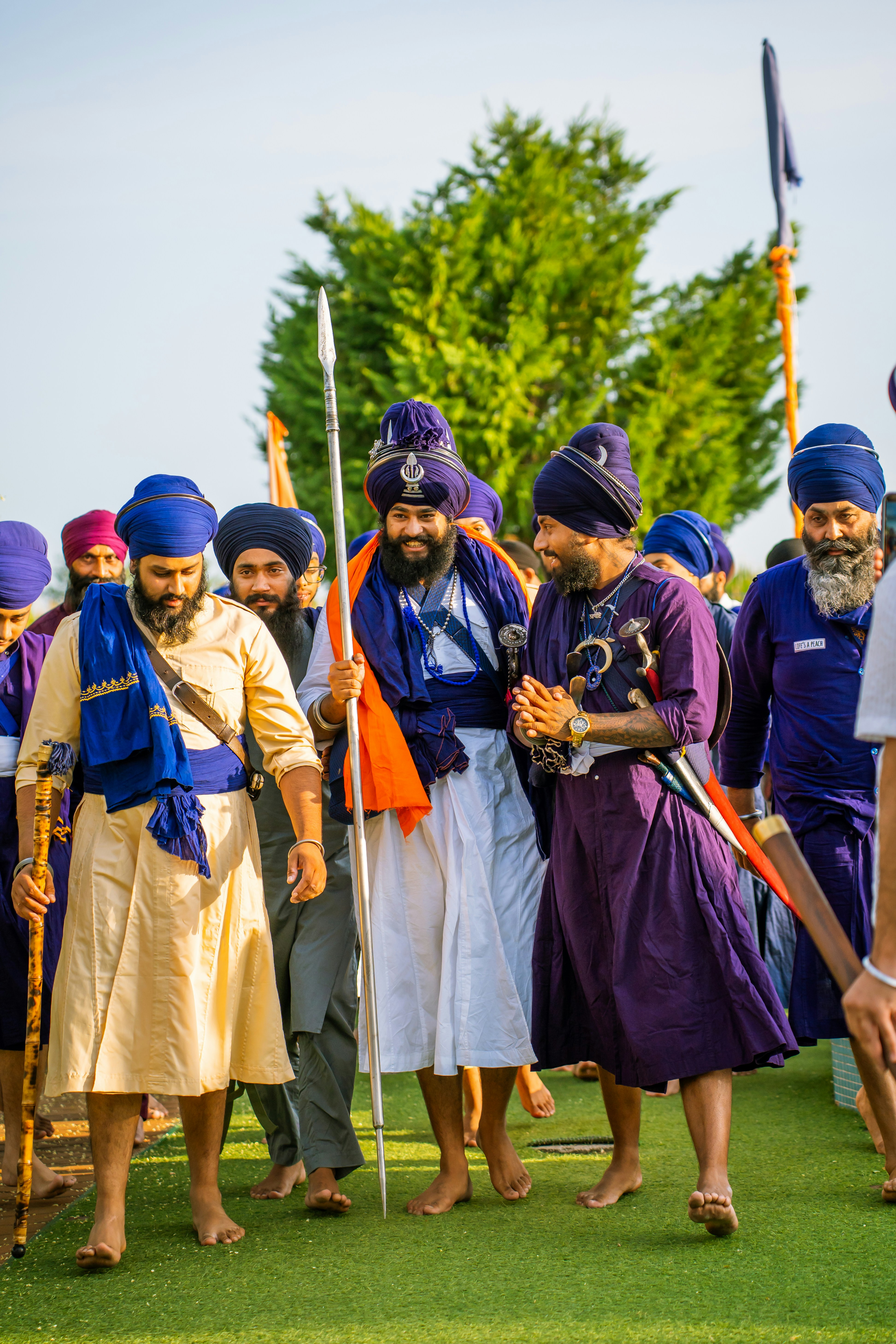 Sikh warriors march in ceremonial attire with spears on a sunlit lawn