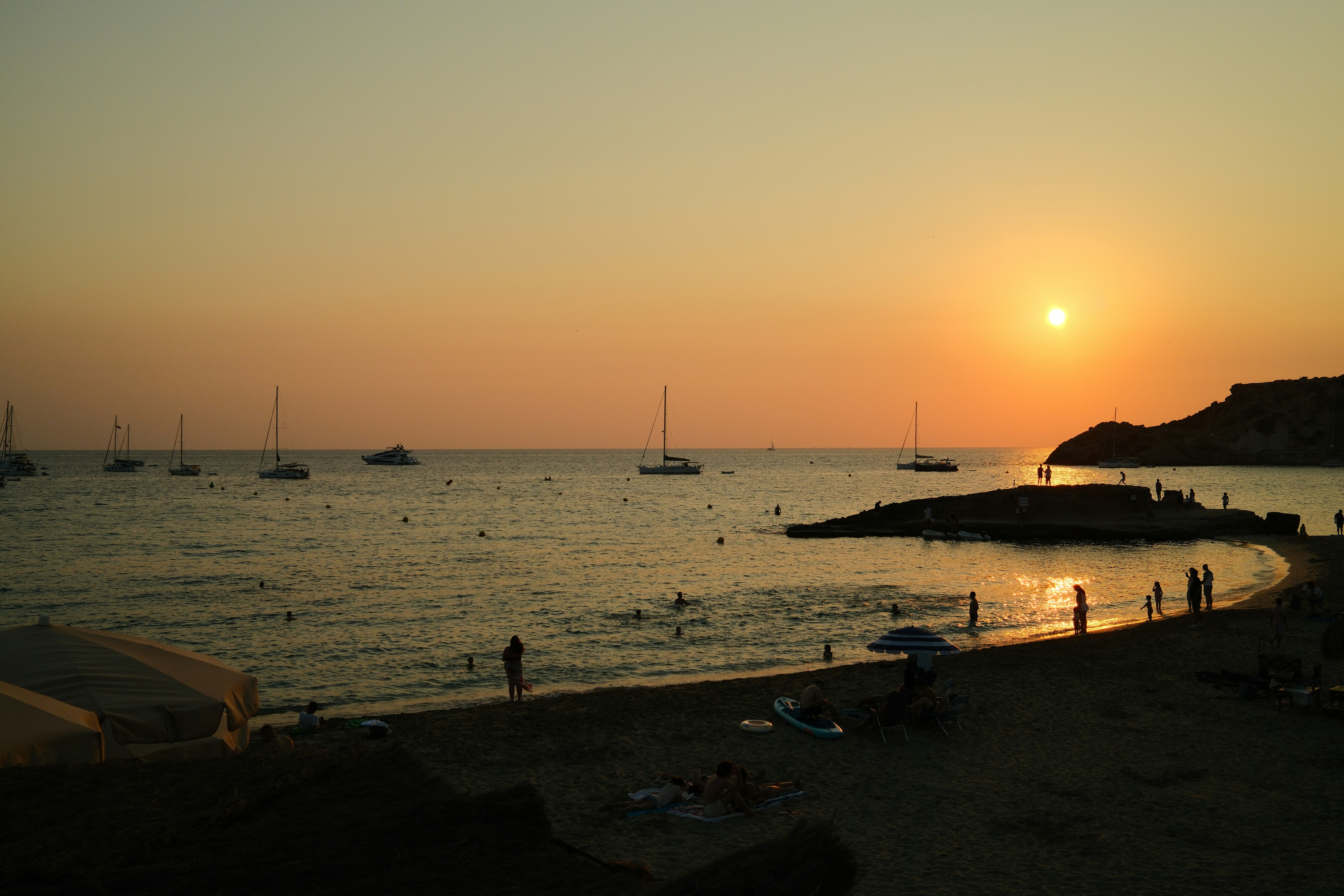 Golden hour at the beach with silhouettes of people and boats along the shore; a calm sea under a warm sky near the sunset.