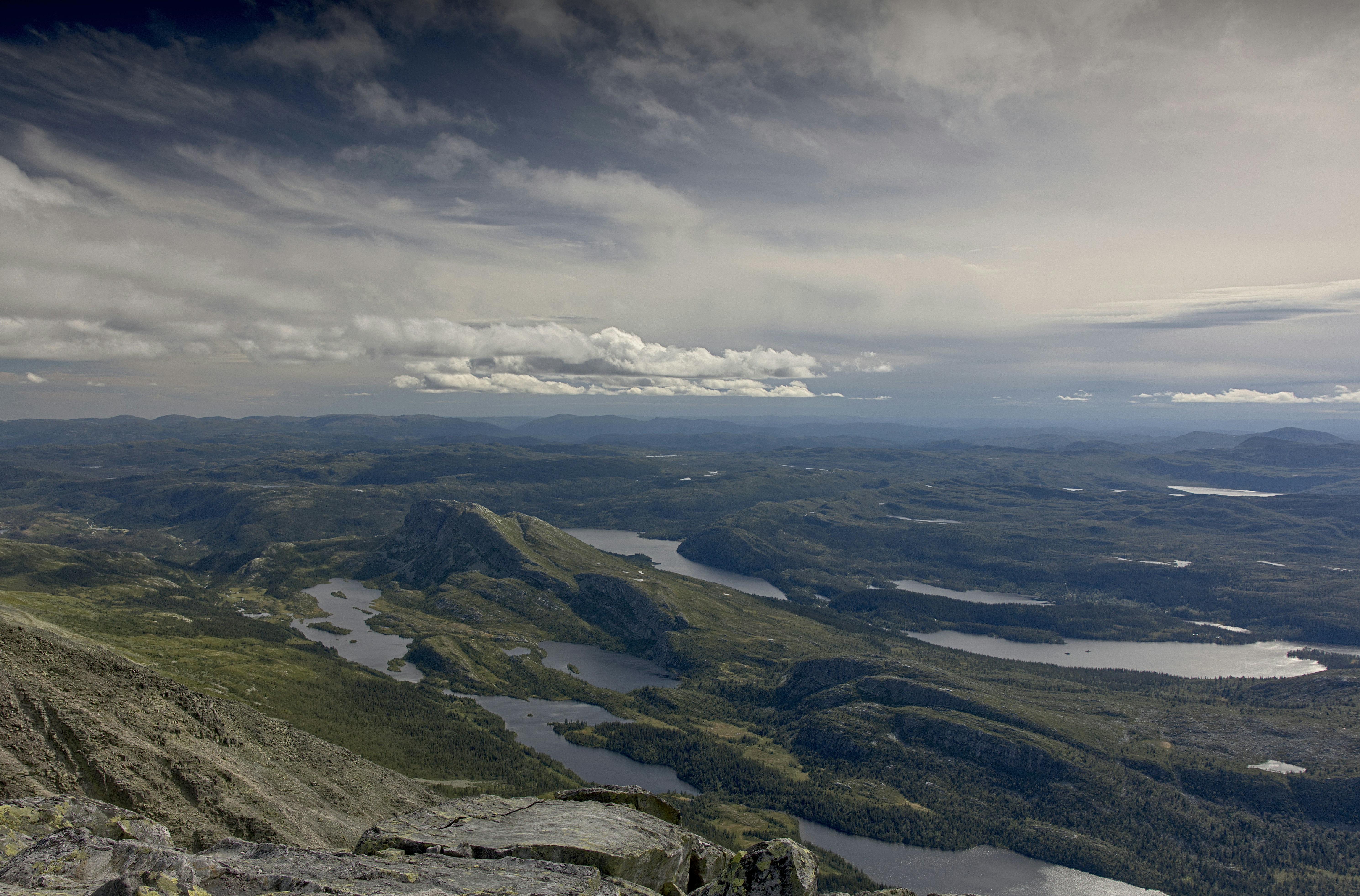 Wide-angle summit panorama revealing rugged foreground ledges, rolling valleys, and winding rivers beneath a broad sky. A landscape shot emphasizing scale and distance, with a natural landscape scene.