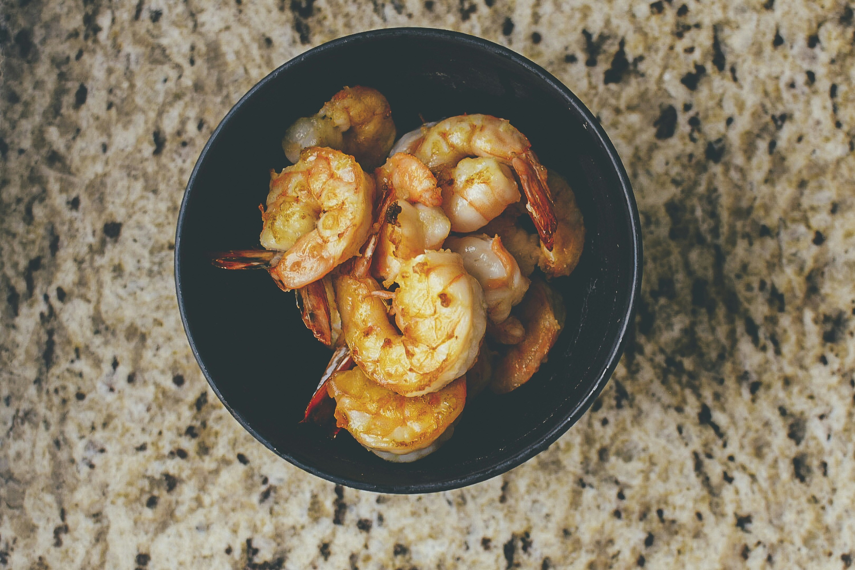 Bright shrimp nest in a dark ceramic bowl set on a speckled countertop, highlighting their orange-pink shells and curled shapes. A culinary close-up showing texture and color detail.