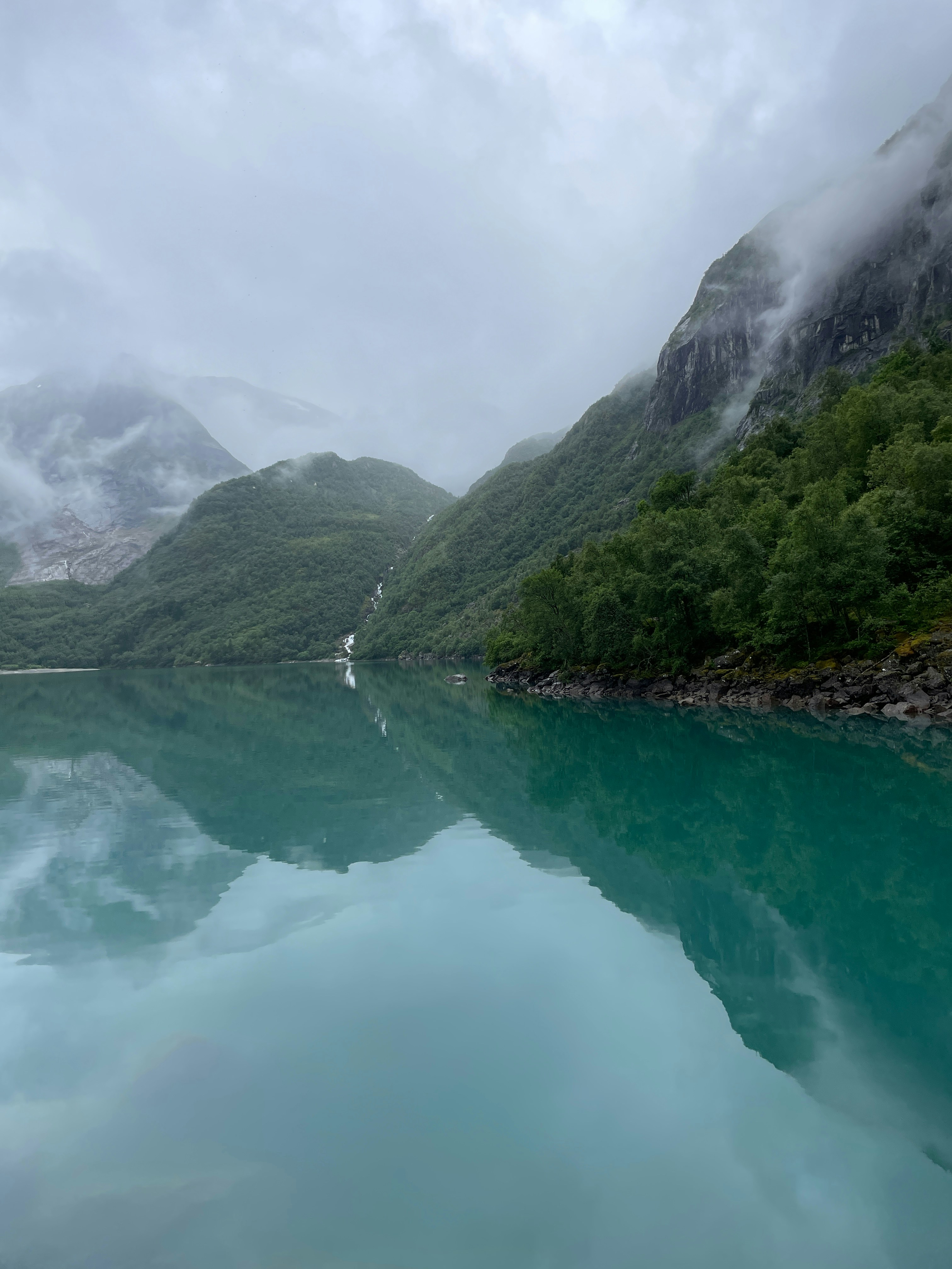 Mist-draped valley with turquoise alpine lake bordered by forested slopes; distant cascading stream and rugged peaks reflected on glassy surface. A tranquil alpine landscape captured in soft, moody light. (Note: photograph of a natural landscape, not a logo or illustration.)