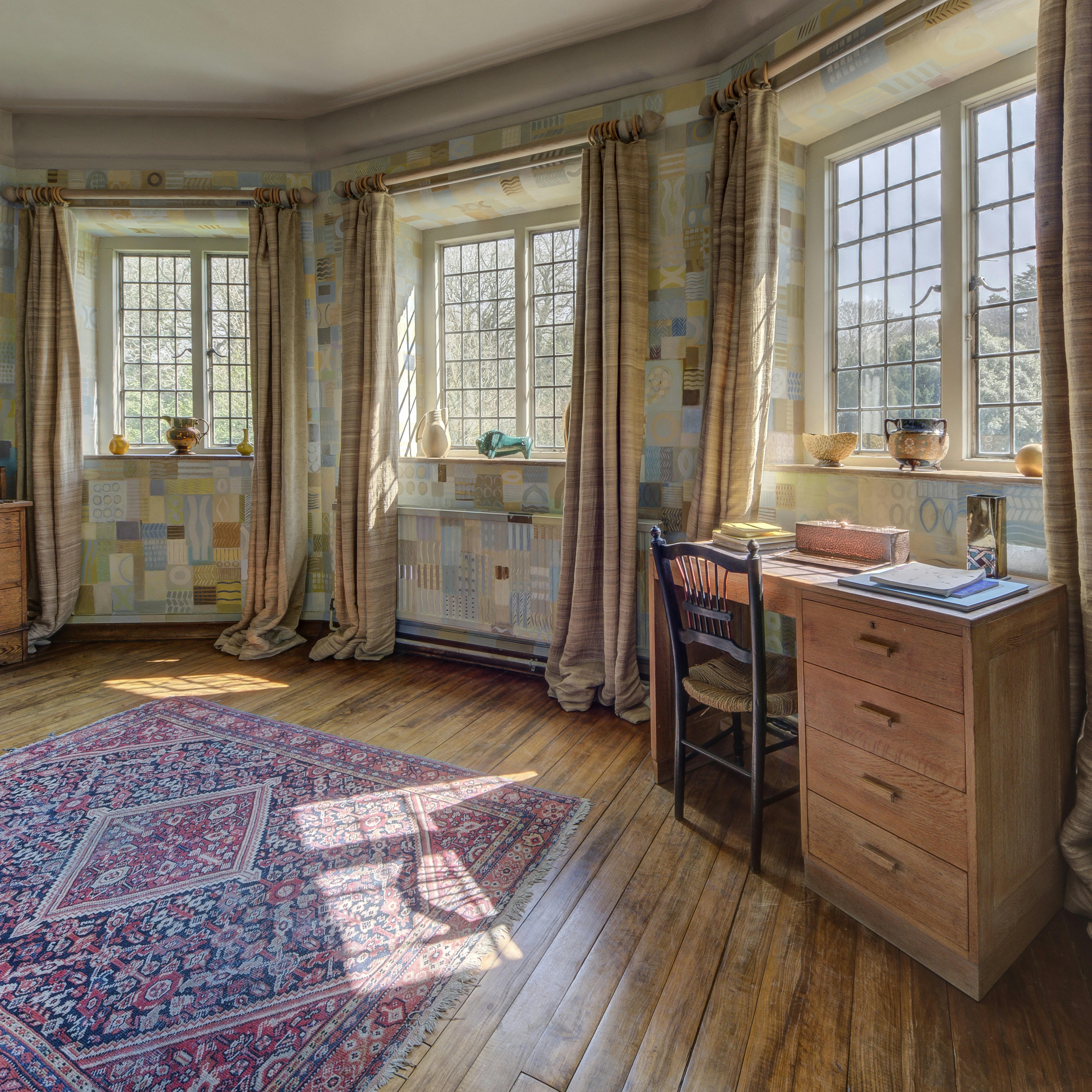 Warmly lit study with patchwork wallpaper, wooden desk and chair by a sunlit window; a patterned rug anchors the oak floor as soft curtains frame the scene.